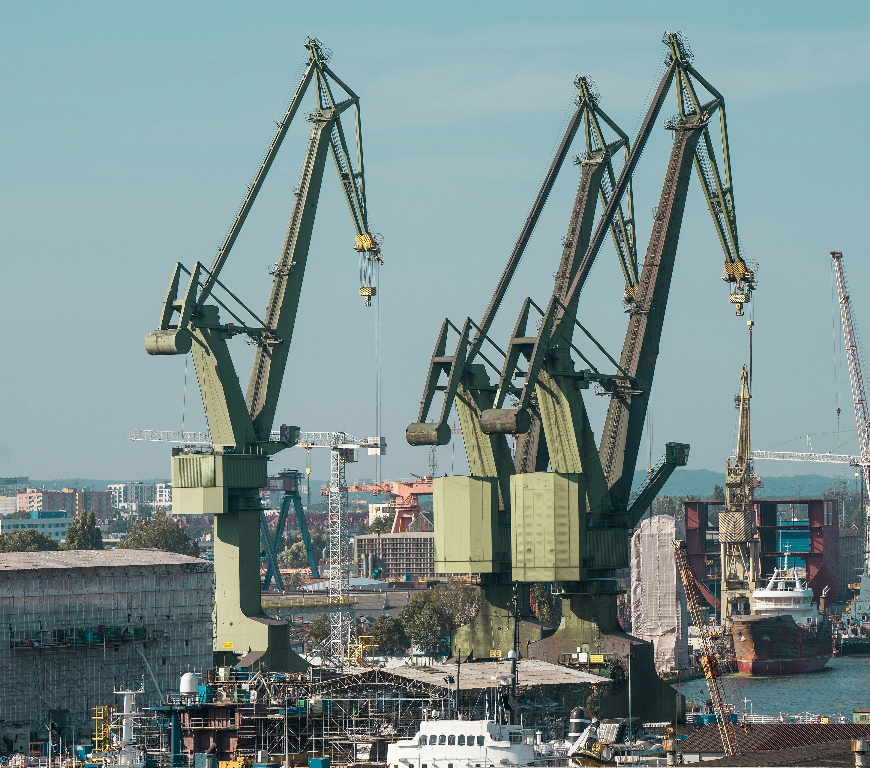 Industrial harbor photograph featuring towering green gantry cranes over the water with docked ships, set against a distant city skyline.