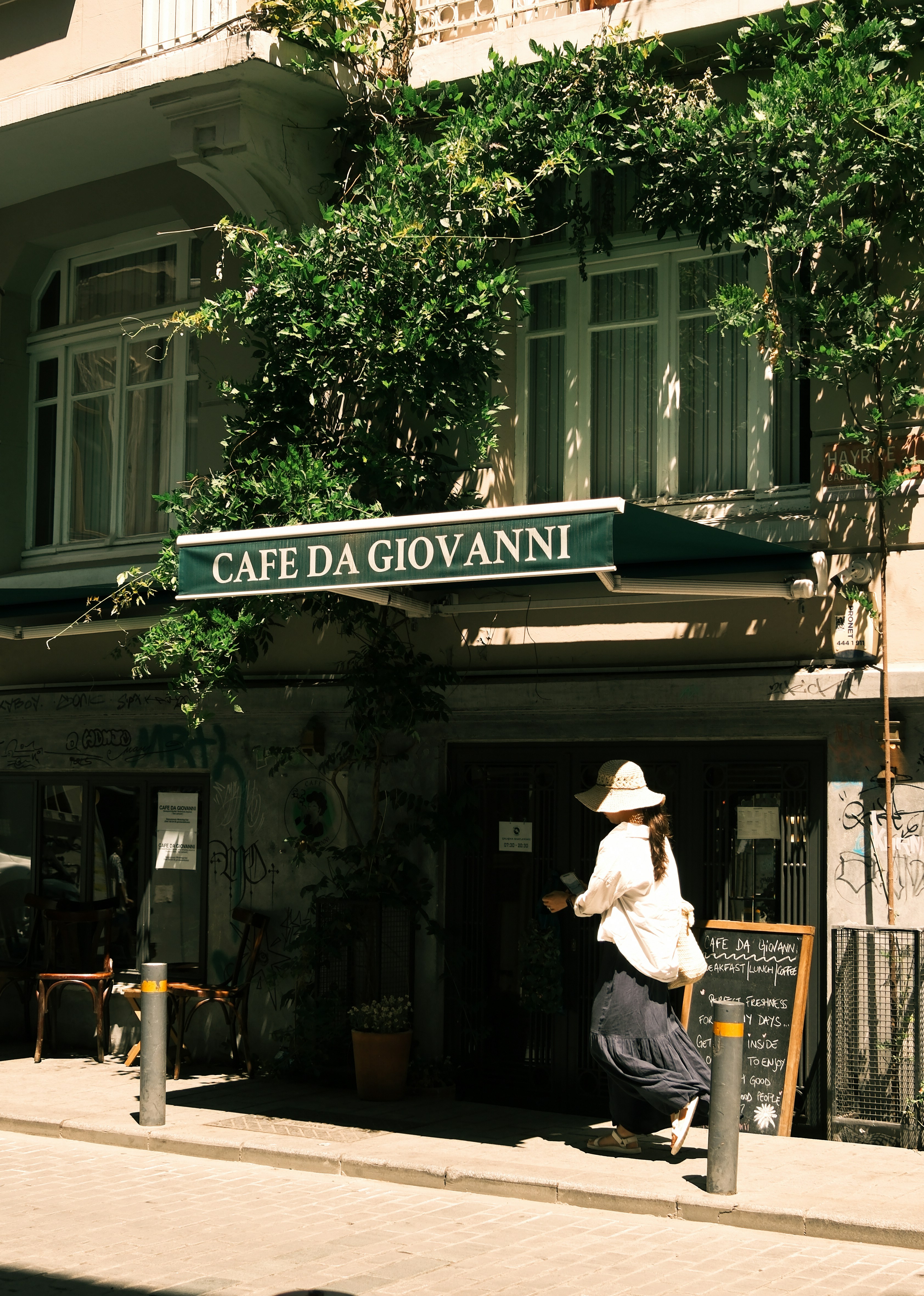 A sunlit street scene with a woman in a wide-brim hat strolling past the green awning of Cafe Da Giovanni. Lush vines frame the storefront, adding natural contrast to the urban café setting.