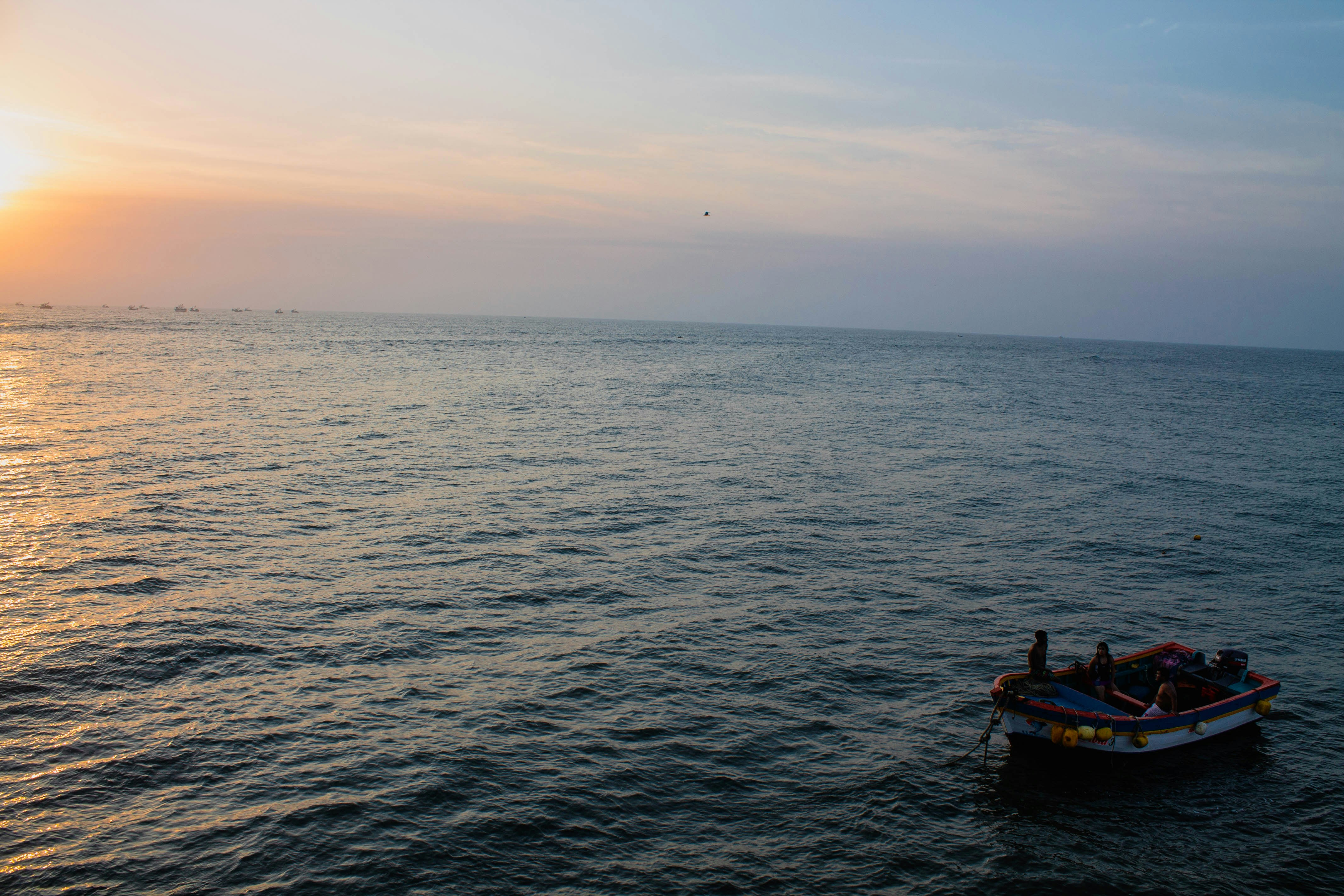 Small boat drifts on a vast ocean under a sunset sky, casting a warm glow over calm waters.