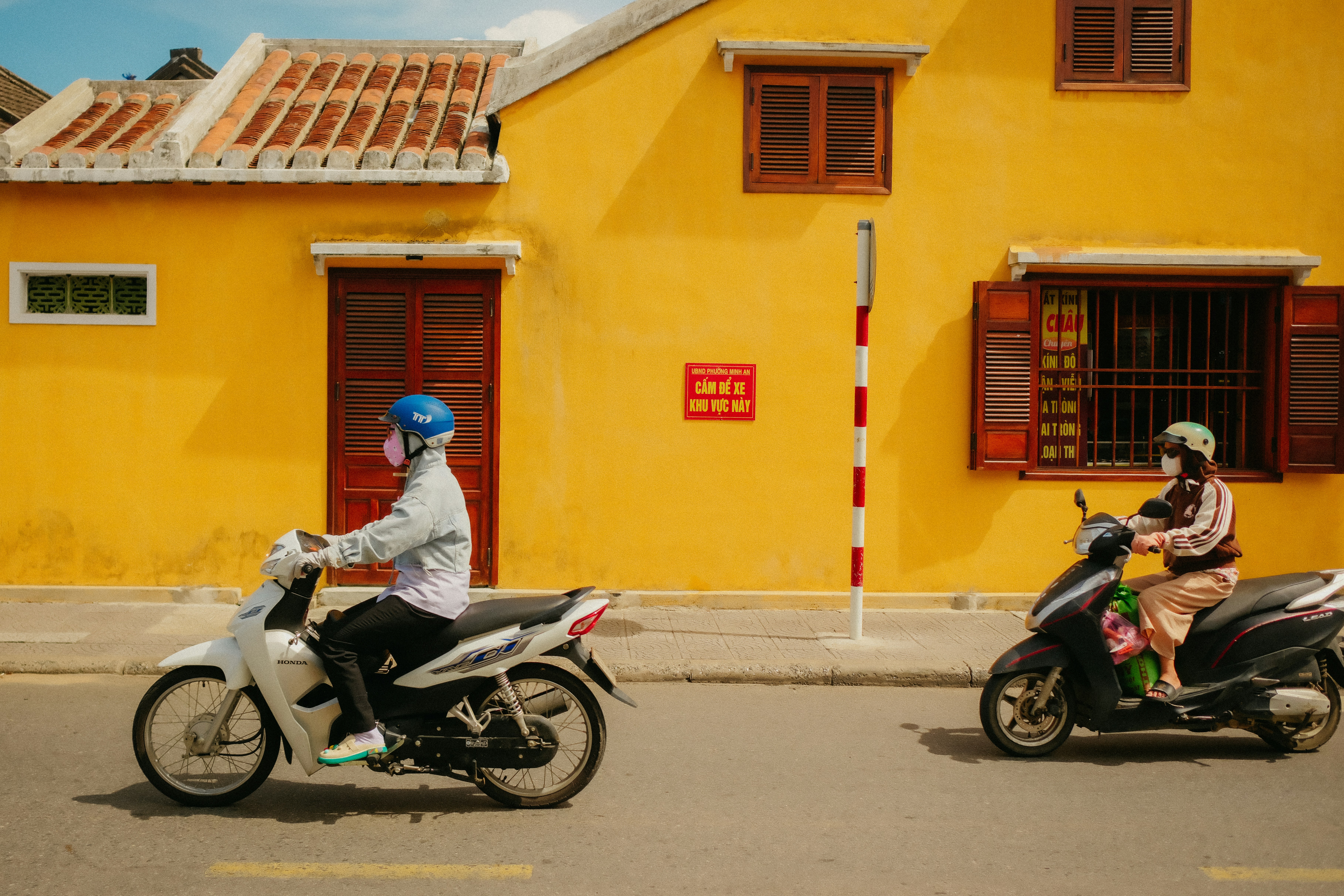 Blue-Helmet Momentum: Scooters Pass a Sunlit Yellow Facade
