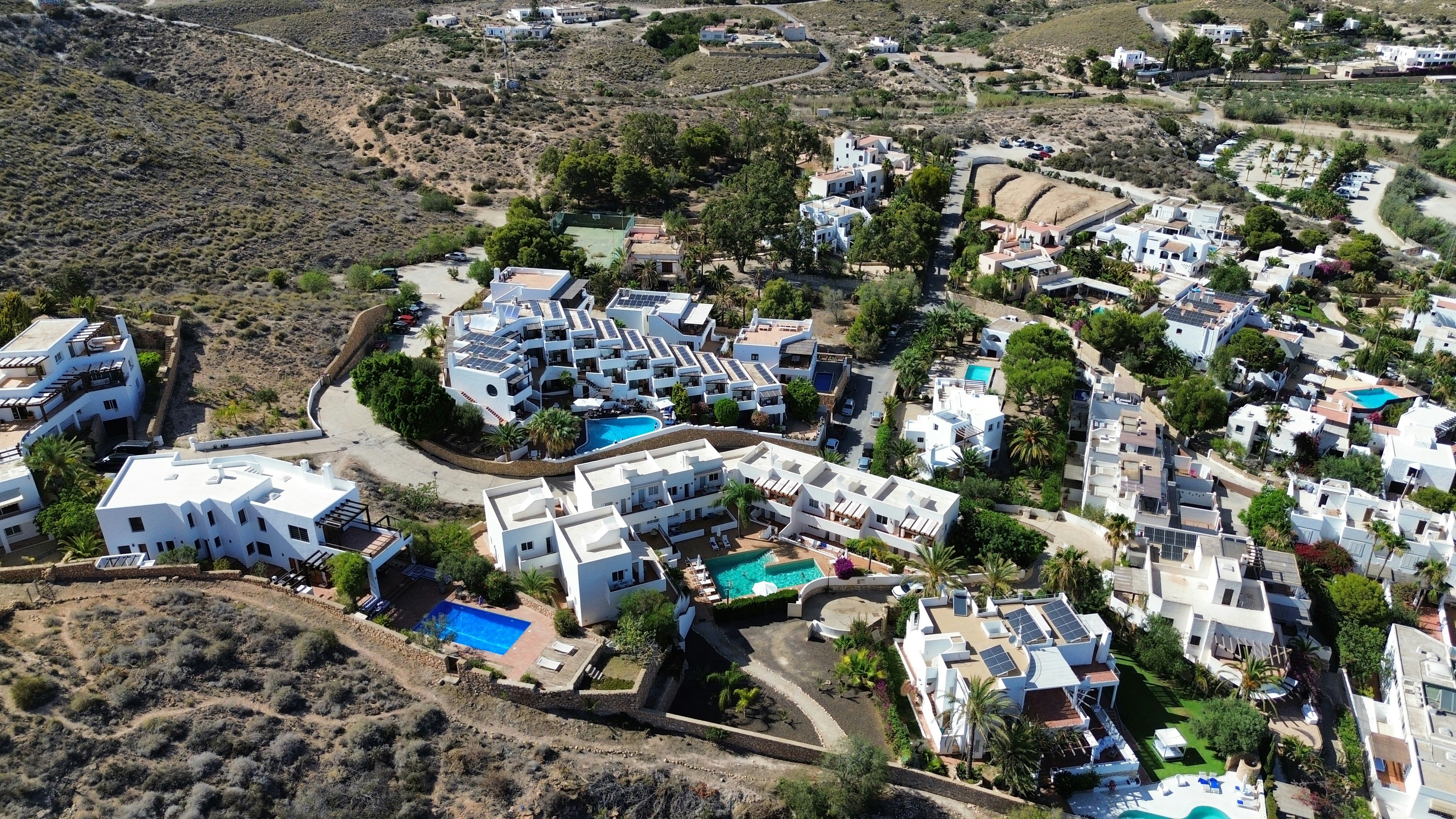 Cluster of white villas with blue pools nestled in a hilly, green landscape.