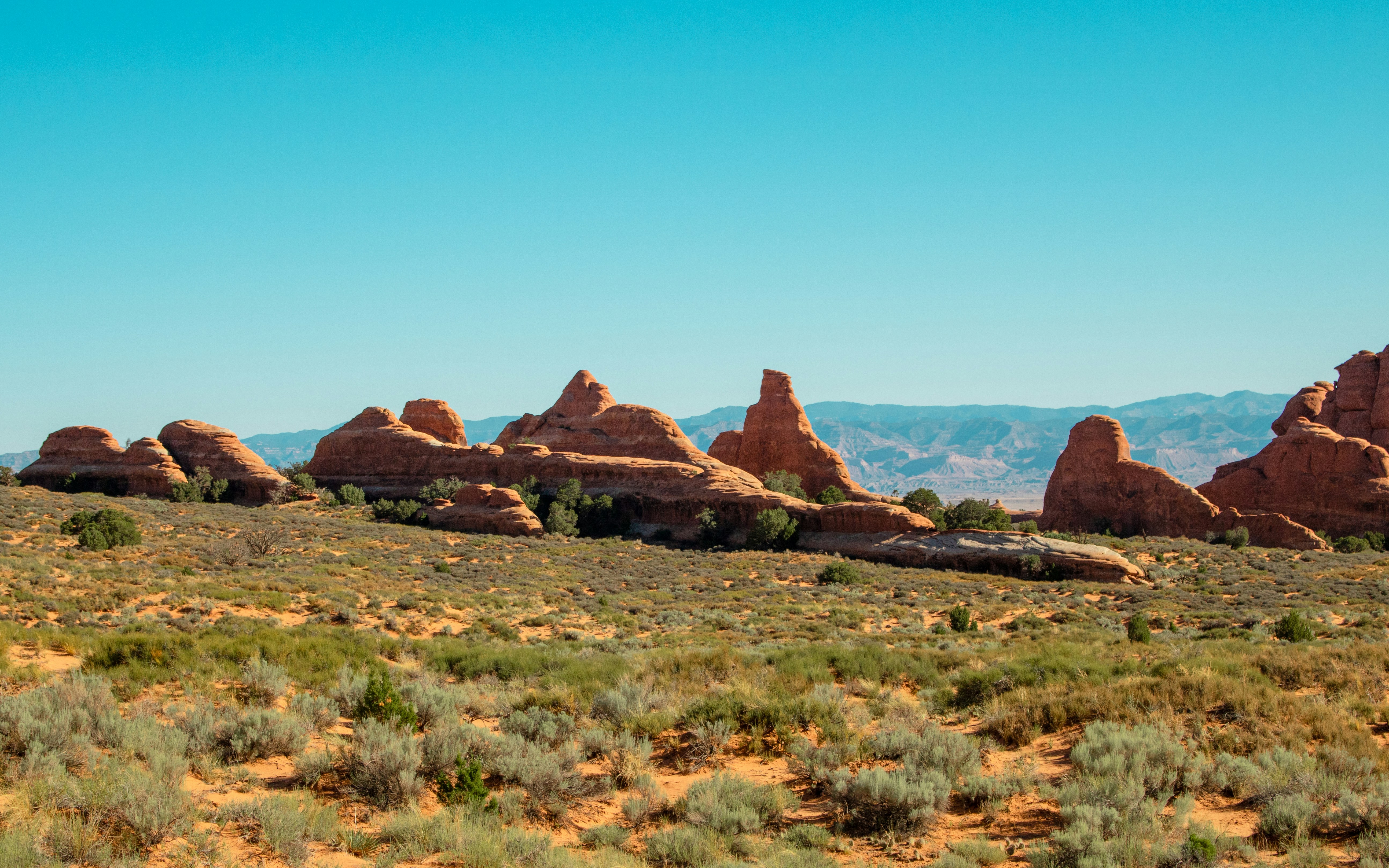 Unique sandstone formations rise from the desert floor, framed by a clear blue sky and distant mountains. The landscape showcases the rugged beauty of nature's artistry.