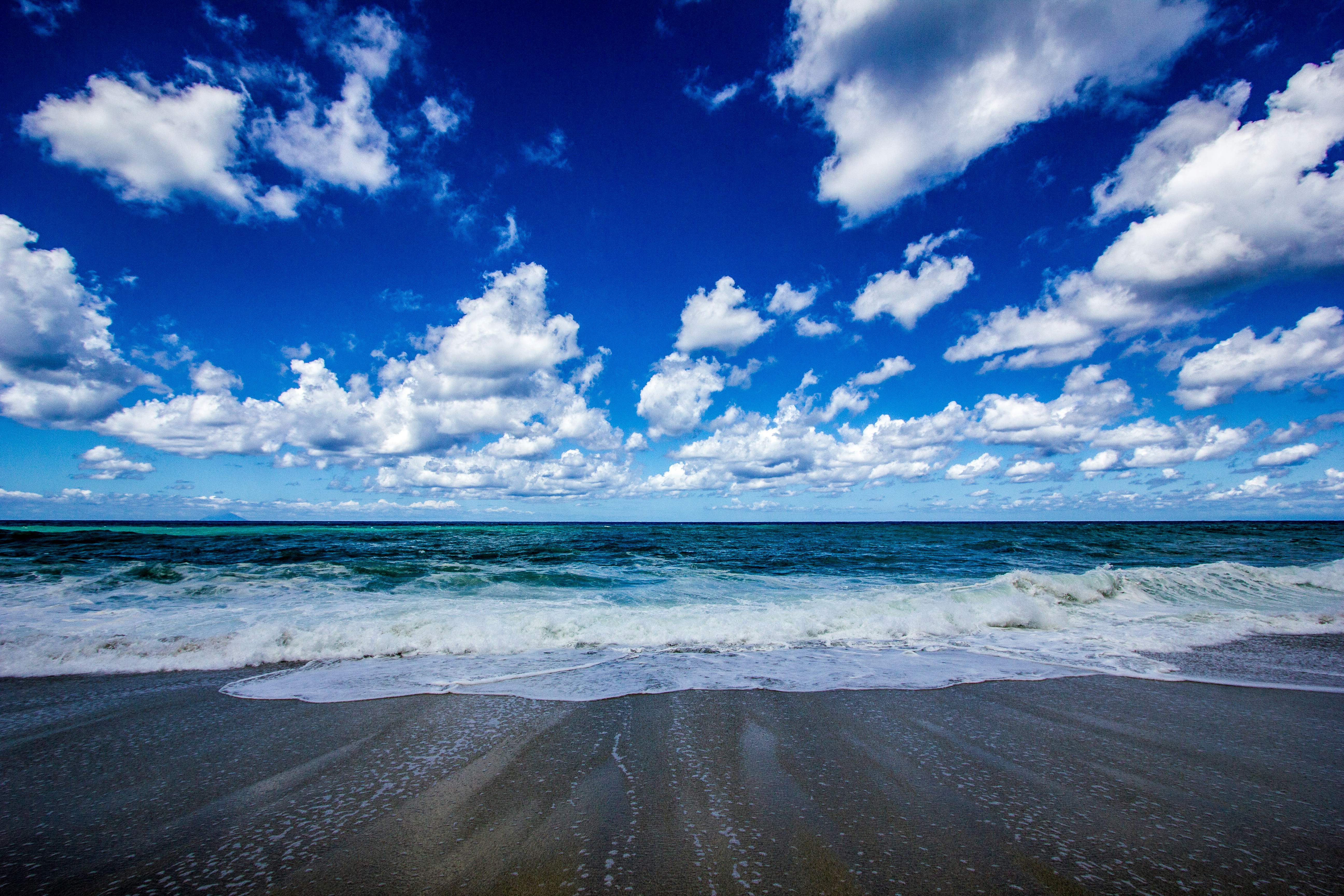 Vibrant blue sky with scattered clouds over a dynamic seascape and gentle waves meeting the sandy shore.