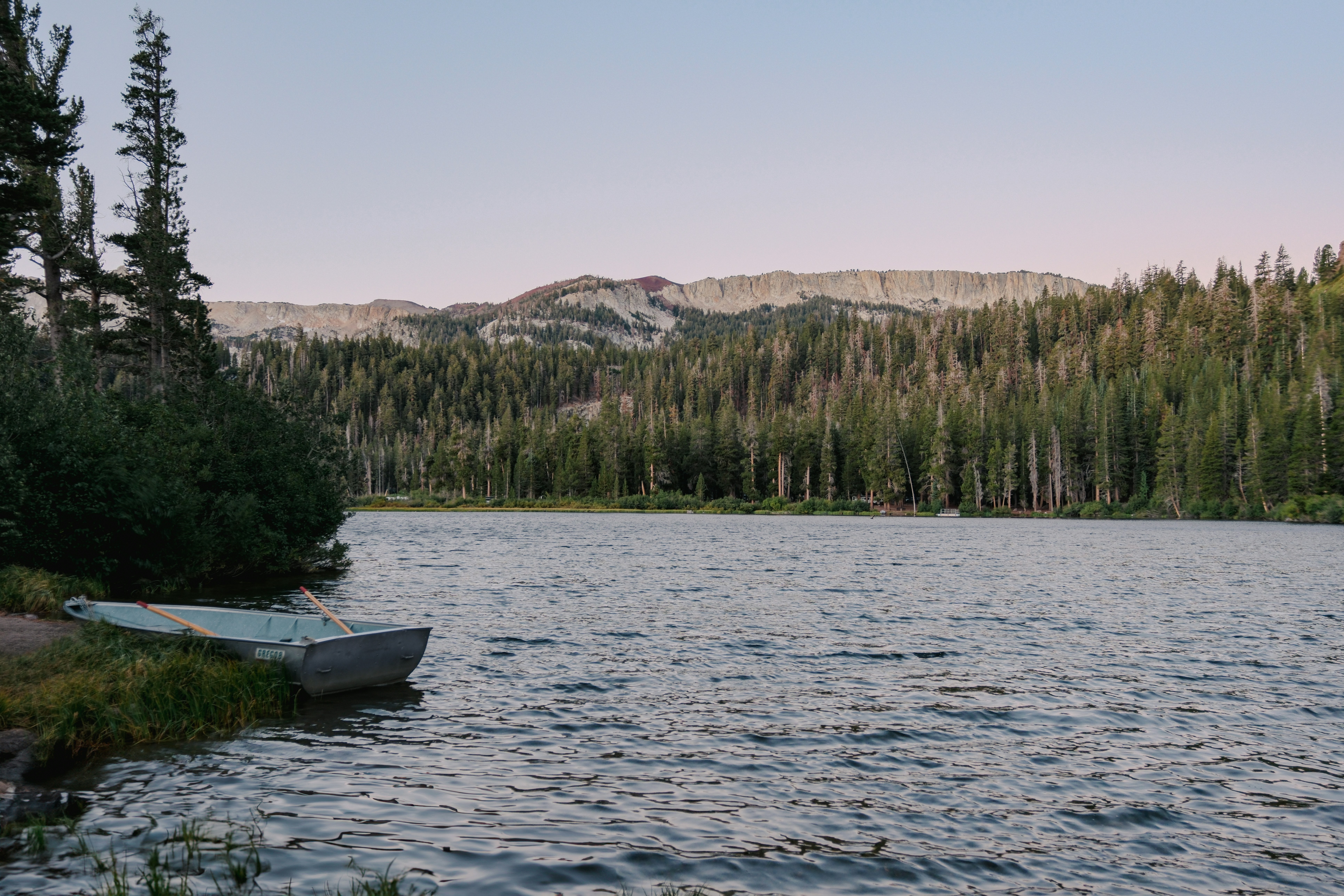 A quiet lakeside scene featuring a small boat resting on the shore, surrounded by lush trees and distant mountains. The calm water reflects the serene atmosphere.