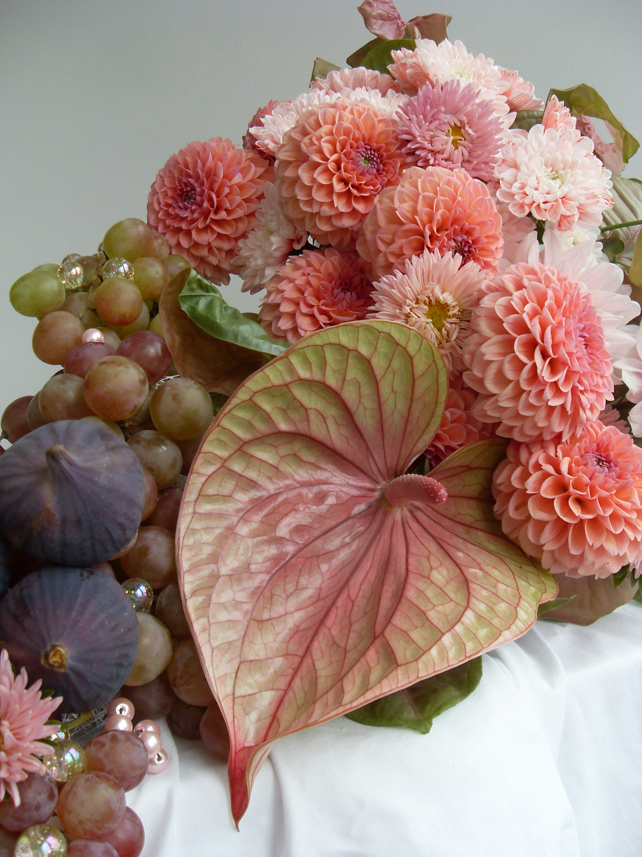 This image creates a dreamy, ethereal atmosphere. In the foreground, there is a stunning arrangement of flowers: dahlias, chrysanthemums, and asters, paired with green and reddish-brown leaves. The floral arrangement is complemented by grapes and figs, giving the composition a rich, still-life appearance. The composition is delicate, with soft lighting that enhances the softness of the colors, creating a calm and artistic visual effect.