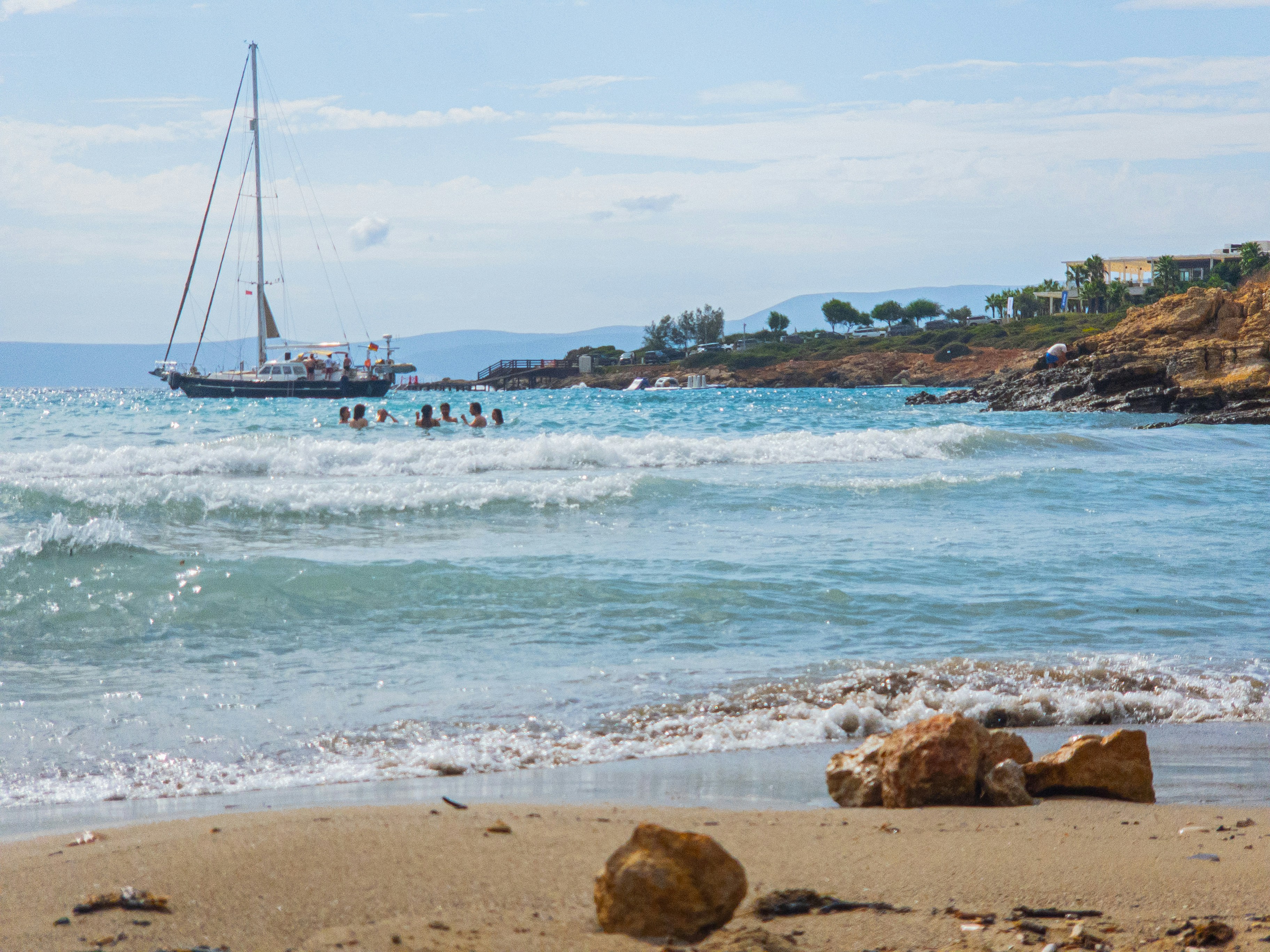 Sailboat near a rocky coastline as turquoise waves break and swimmers gather in the foreground under a bright sky.