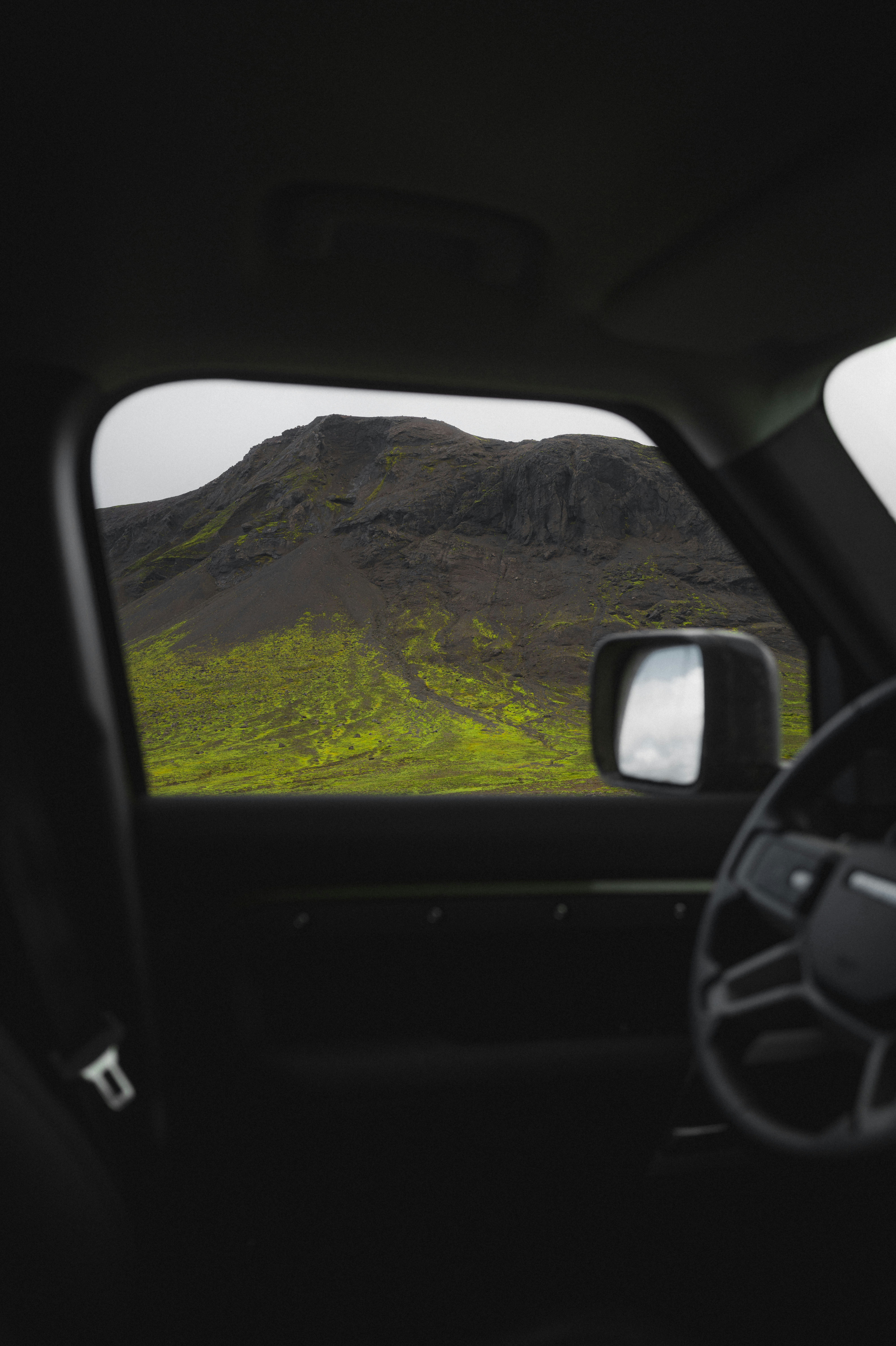 Moss-covered volcanic ridge visible through a car's side window, with the steering wheel and dark interior framing the scene.