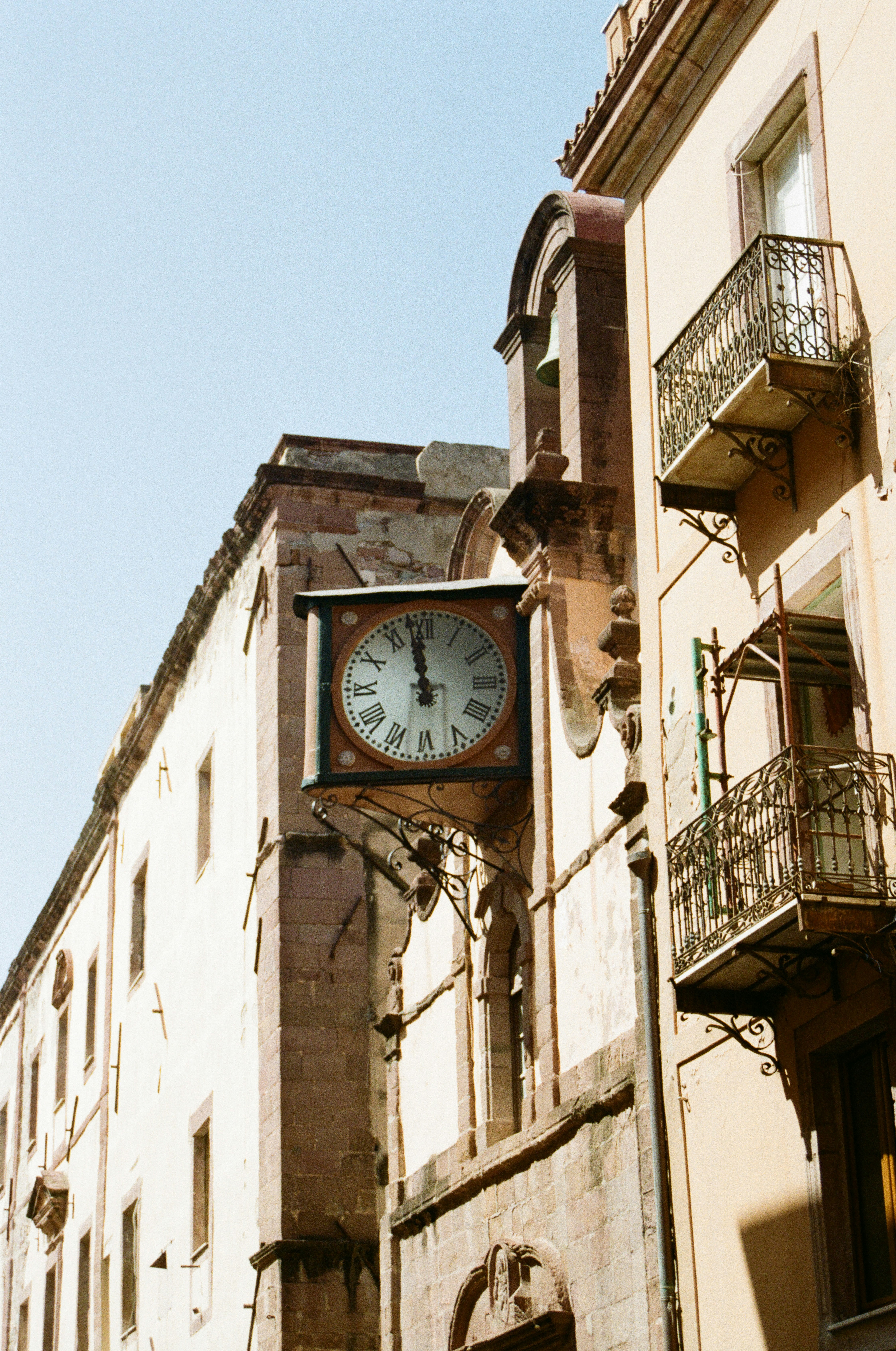 A vintage clock mounted on a weathered stone wall along a sunlit street, with wrought-iron balconies on the adjacent building.