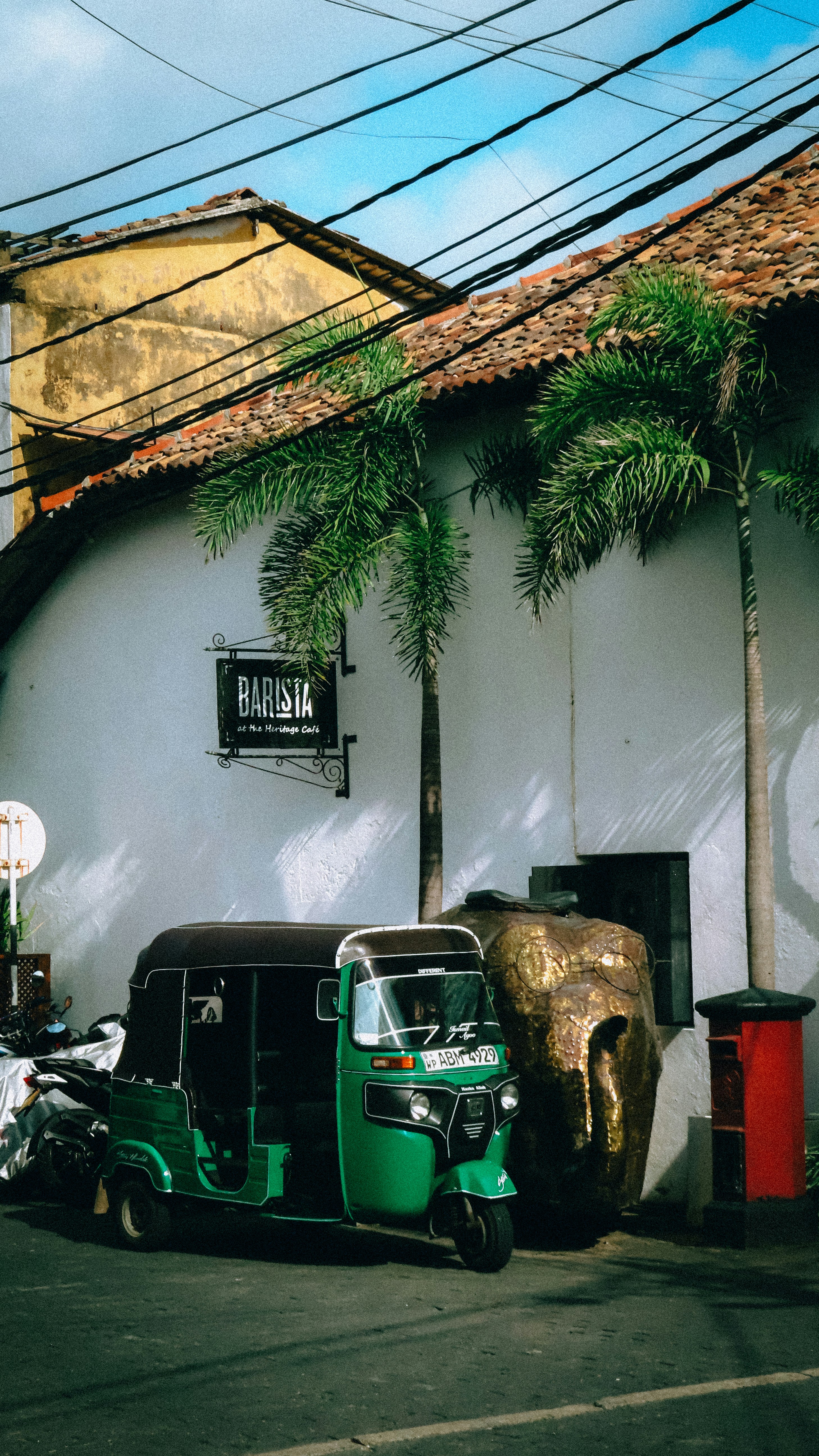 Street photograph of a vivid green auto rickshaw parked beside a white building with a BARISTA sign, beneath overhead cables and palm fronds. The composition emphasizes urban texture and everyday motion.