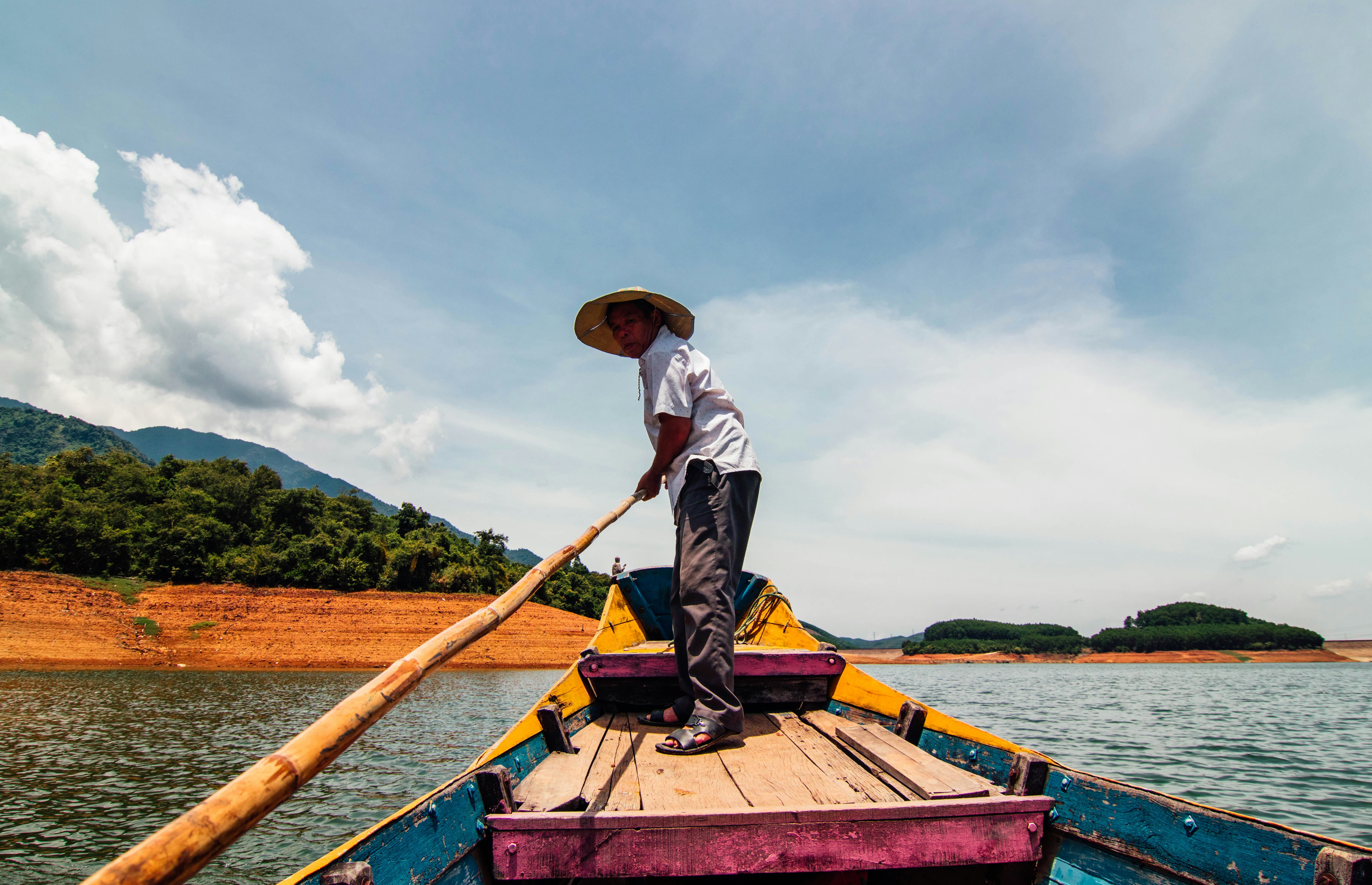 Man rowing a colorful boat on a calm lake with distant green hills under a clear blue sky.