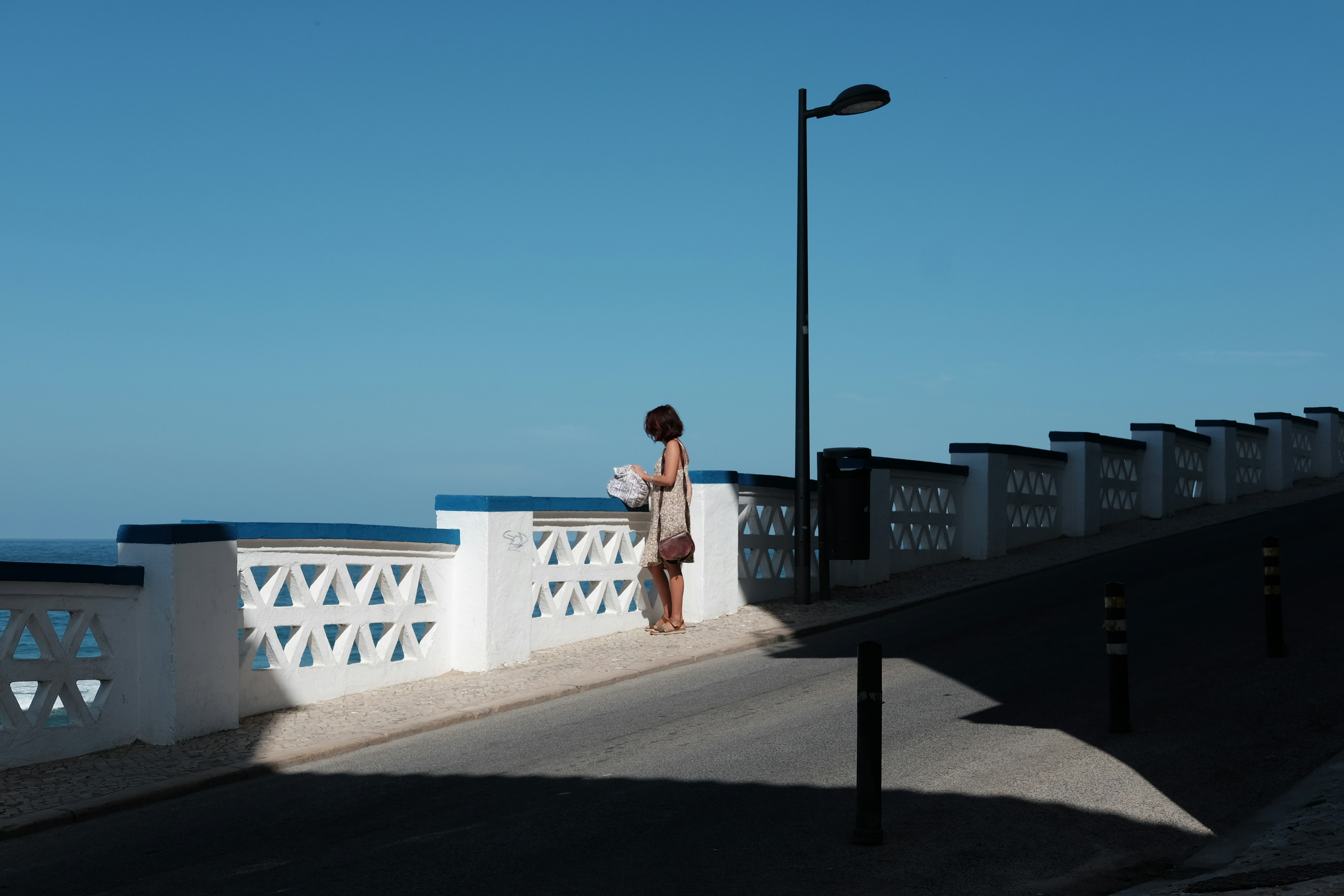 I’m drawn to the way tension holds this frame together, the play between structure and the solitary figure, between stillness and a suggestion of movement. She stands in a hard slice of sunlight, bracketed by the lamppost and the white railing, almost swallowed by the architecture around her. The diagonal pull of the street adds a quiet momentum, while the verticals break up the space rhythmically. It’s that moment where form and human presence coexist in a kind of unresolved balance.