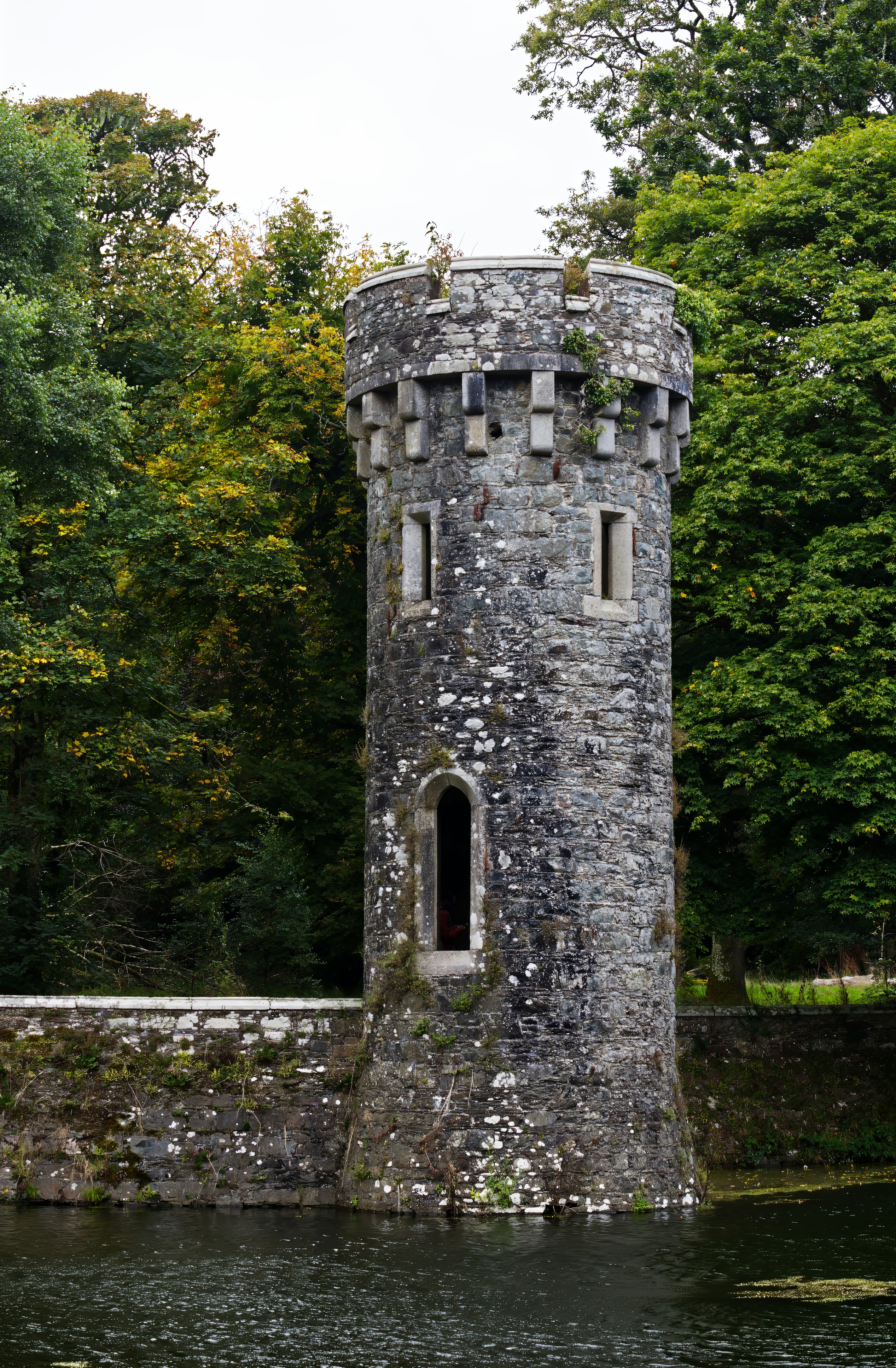 Stone watchtower standing beside a tranquil lake, surrounded by lush greenery.