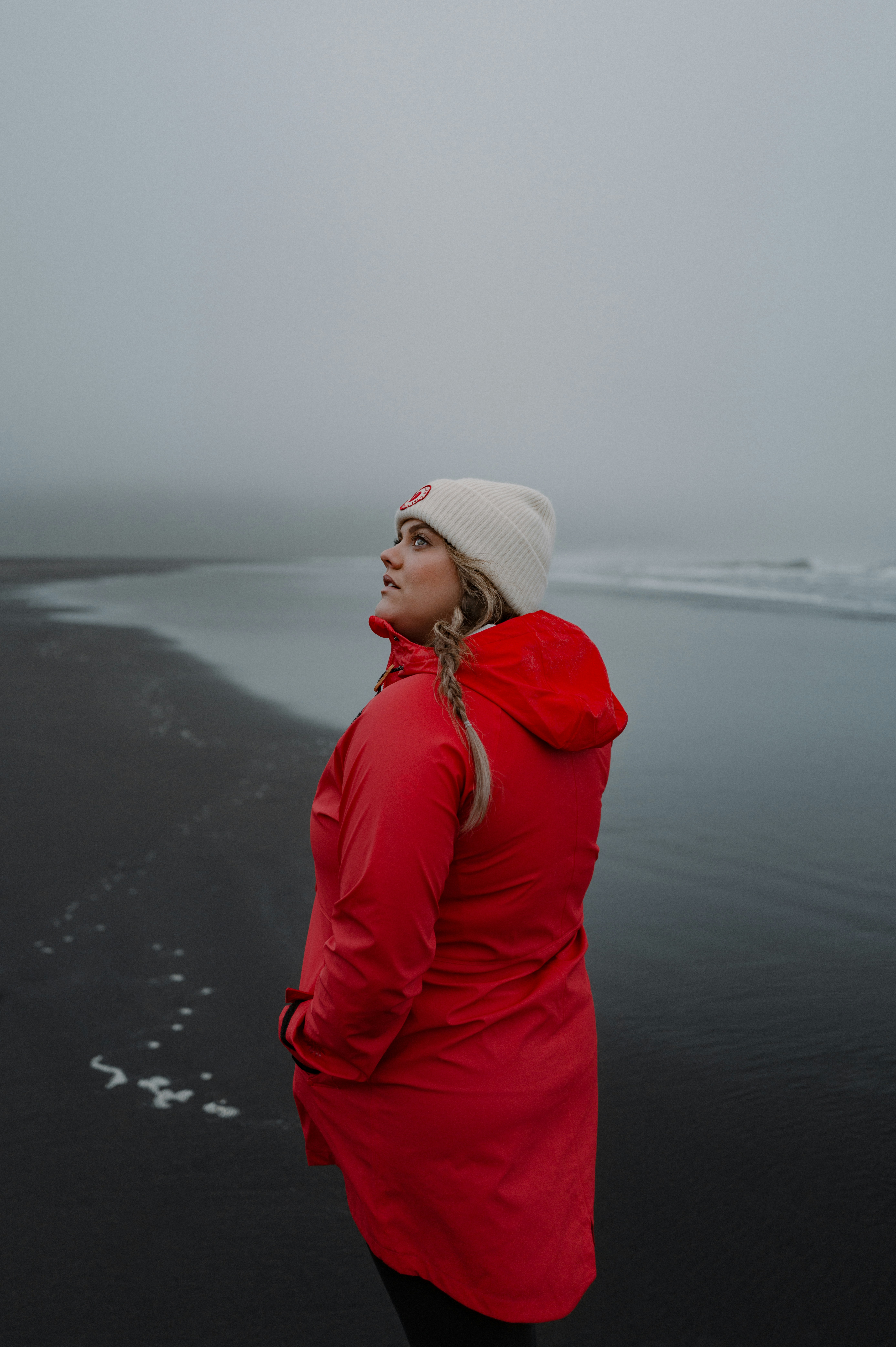 Portrait of a woman in a bright red coat and cream knit hat standing on a dark, misty beach with calm water and an overcast sky.