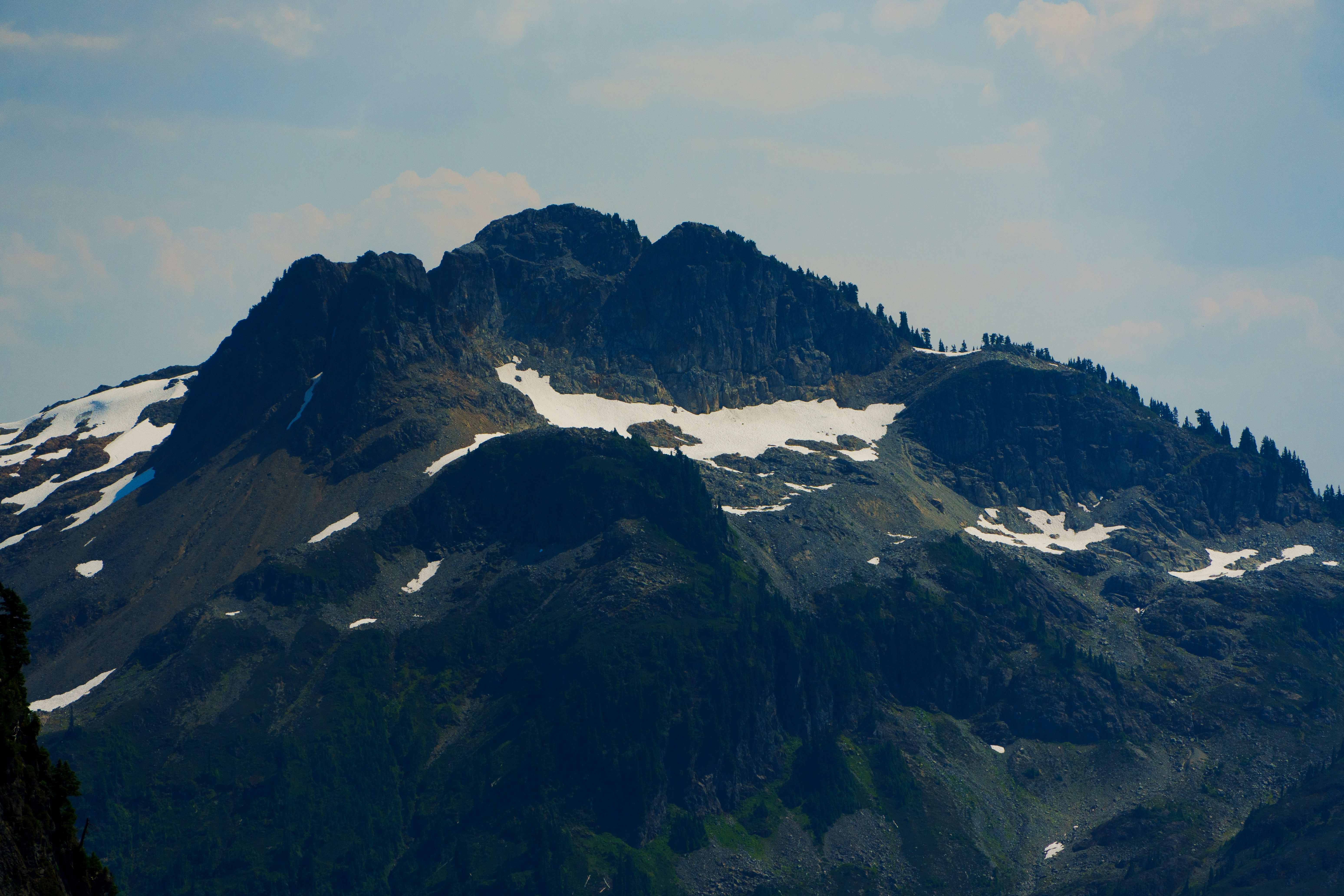 Mountain landscape with patches of snow on dark rocky terrain beneath a lightly clouded sky.