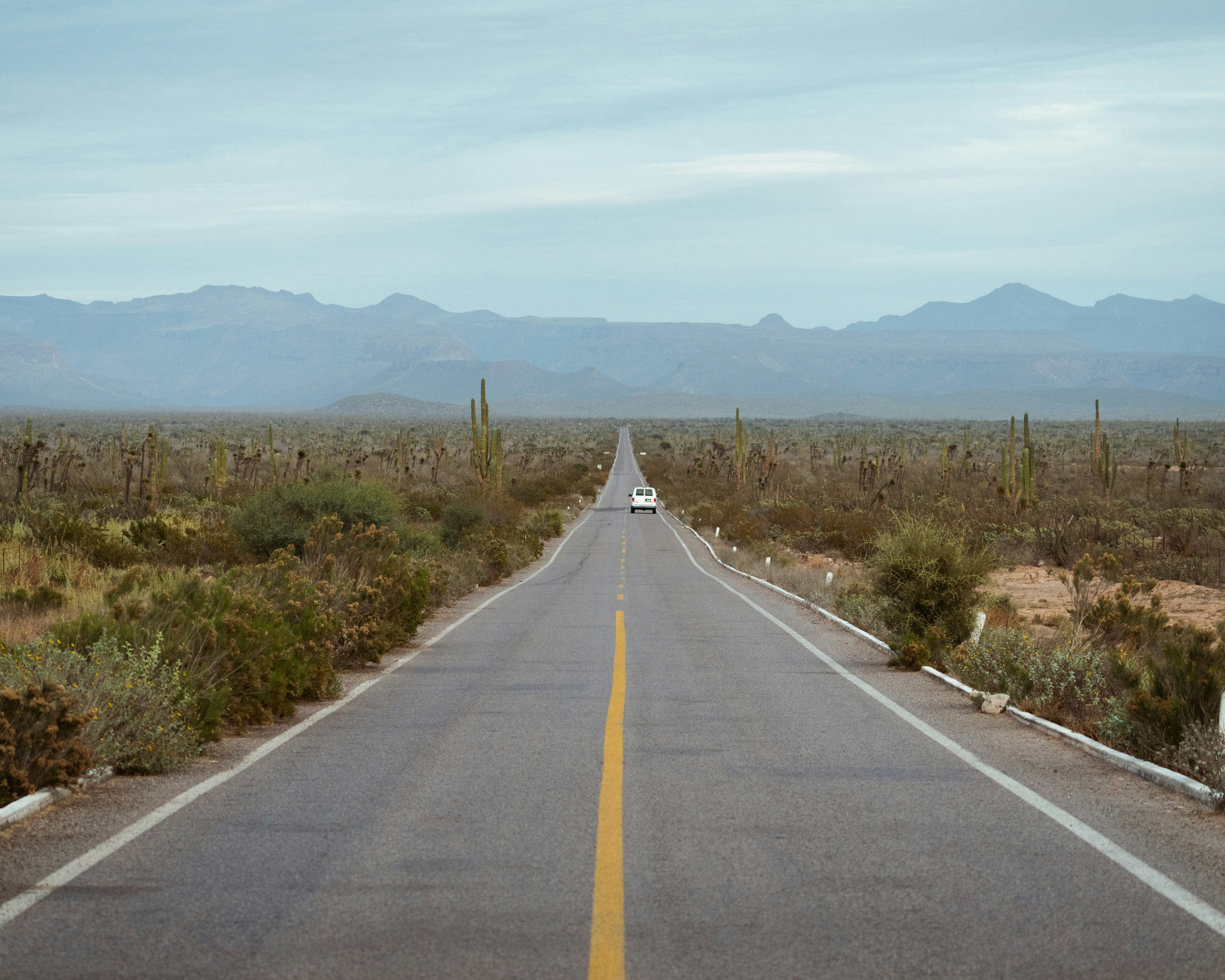 Desert Highway: A Lone White Car Converges Along a Narrow Line Toward Distant Peaks