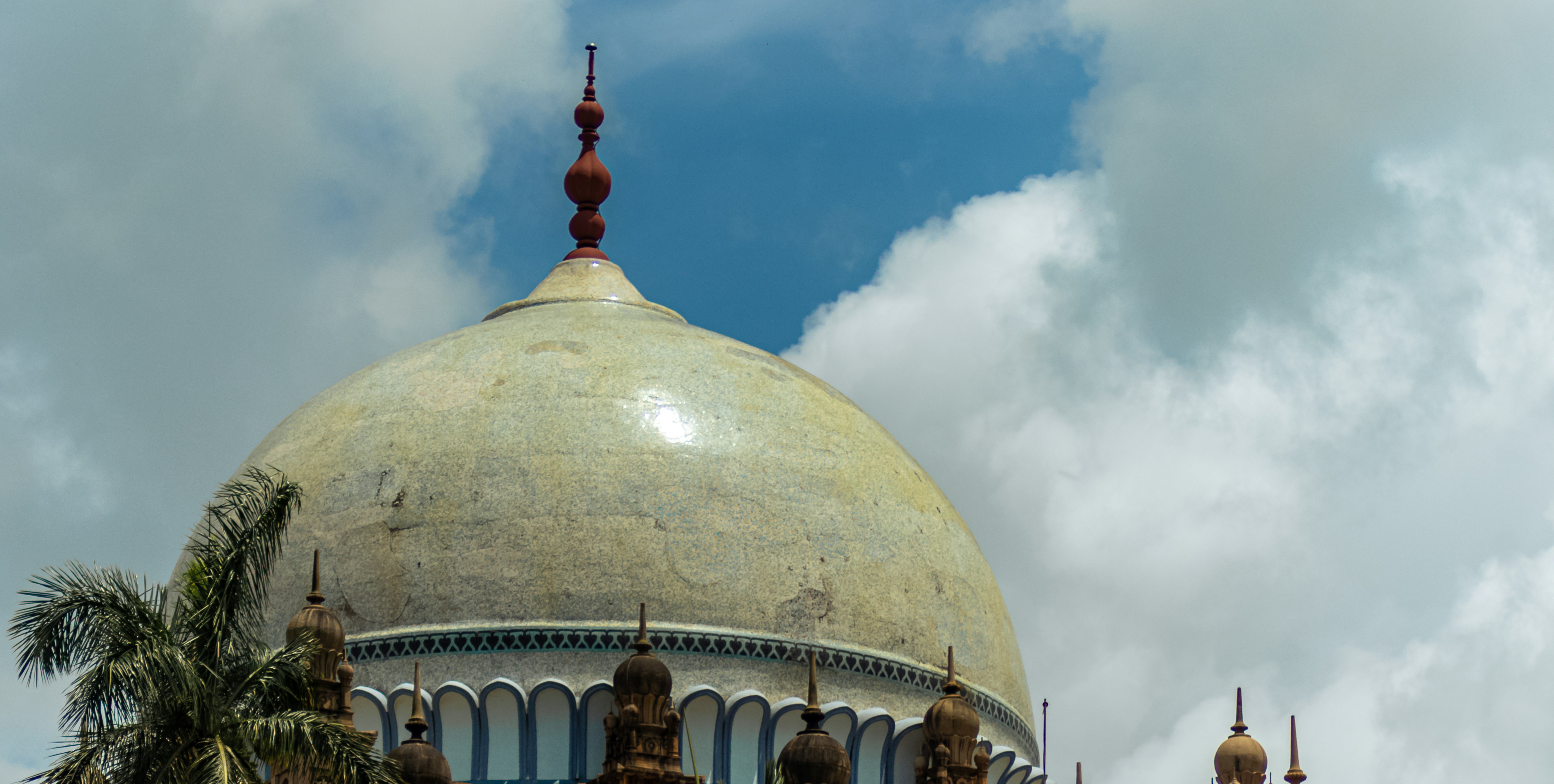 Ornate dome adorned with intricate details, framed by lush palm leaves and a backdrop of dramatic clouds.