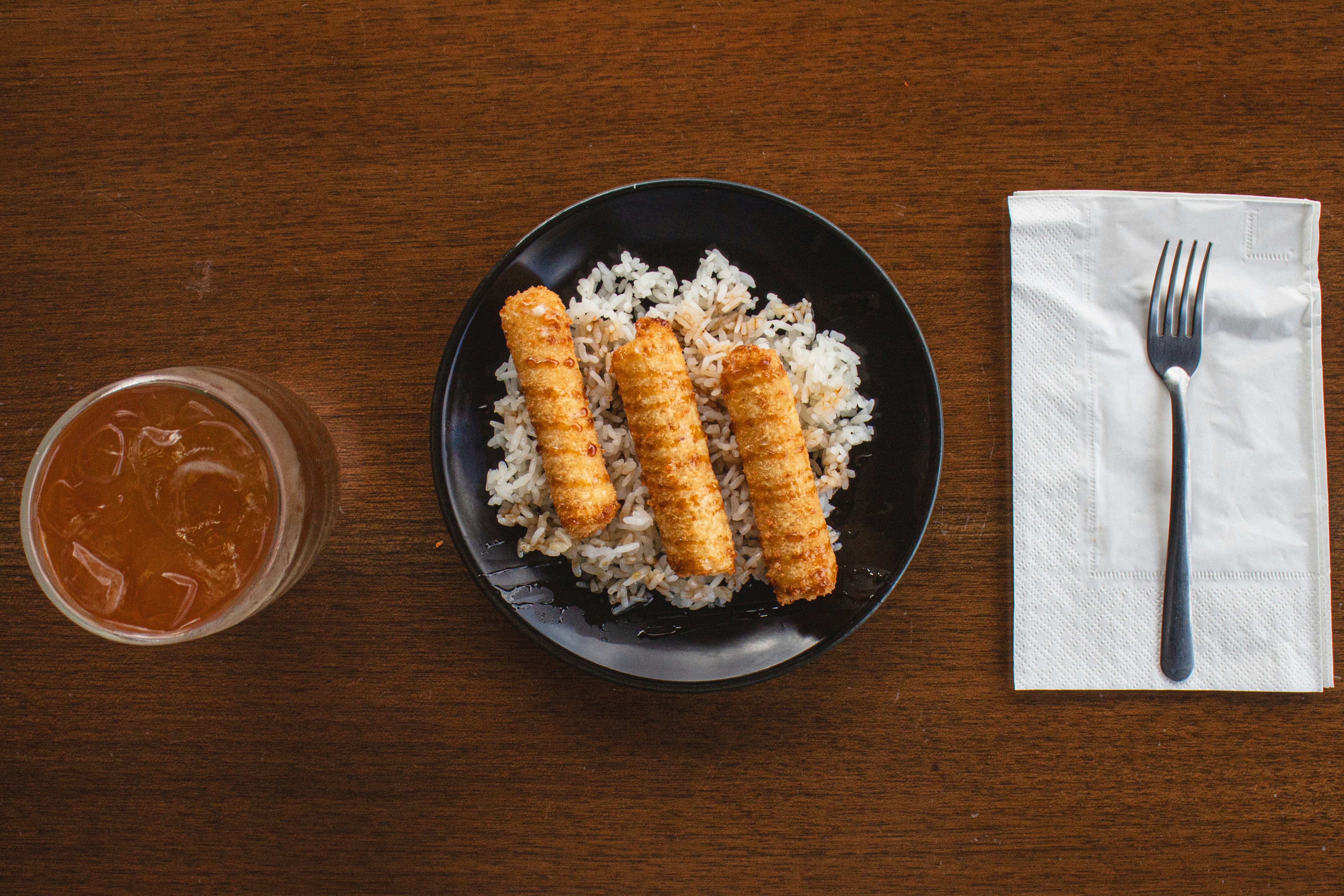 Crispy fish sticks on white rice beside iced tea and a fork on a napkin.