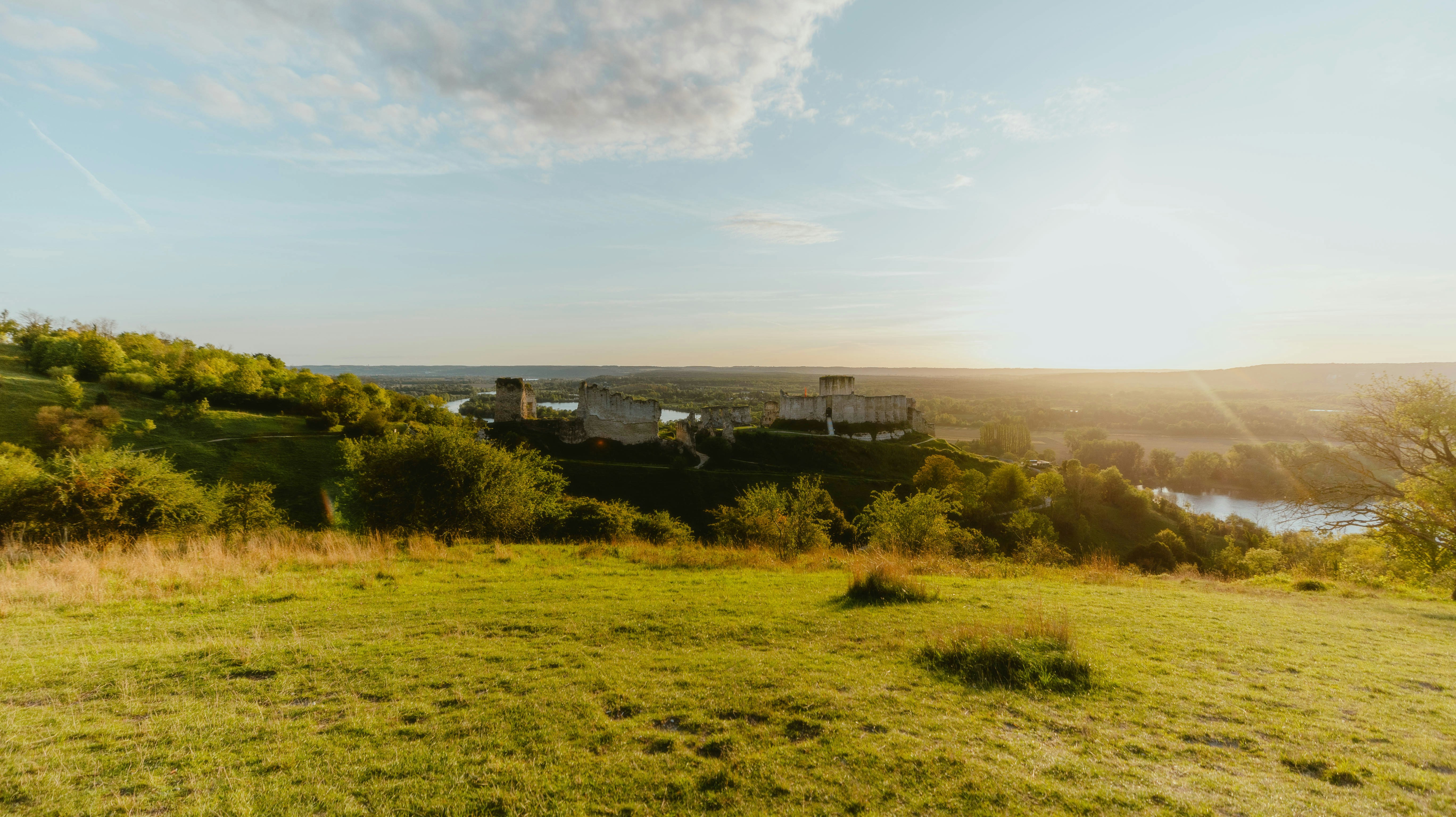 Golden sunlight spills over rolling hills, illuminating a distant structure beside a winding river. The scene captures the tranquil beauty of nature at dusk.
