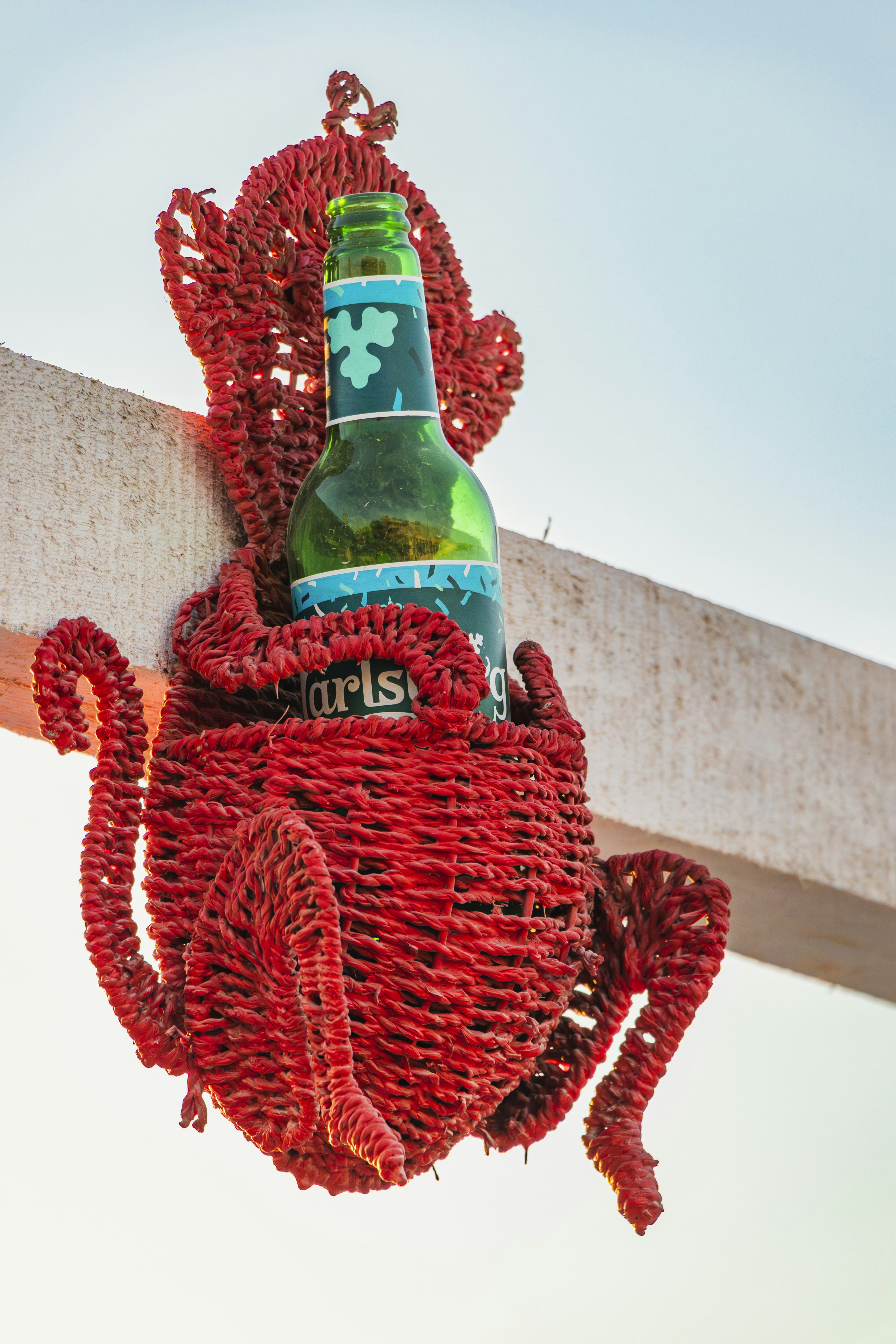 Crimson Weave Cradle: A Beer Bottle Perched on a Sunlit Concrete Beam