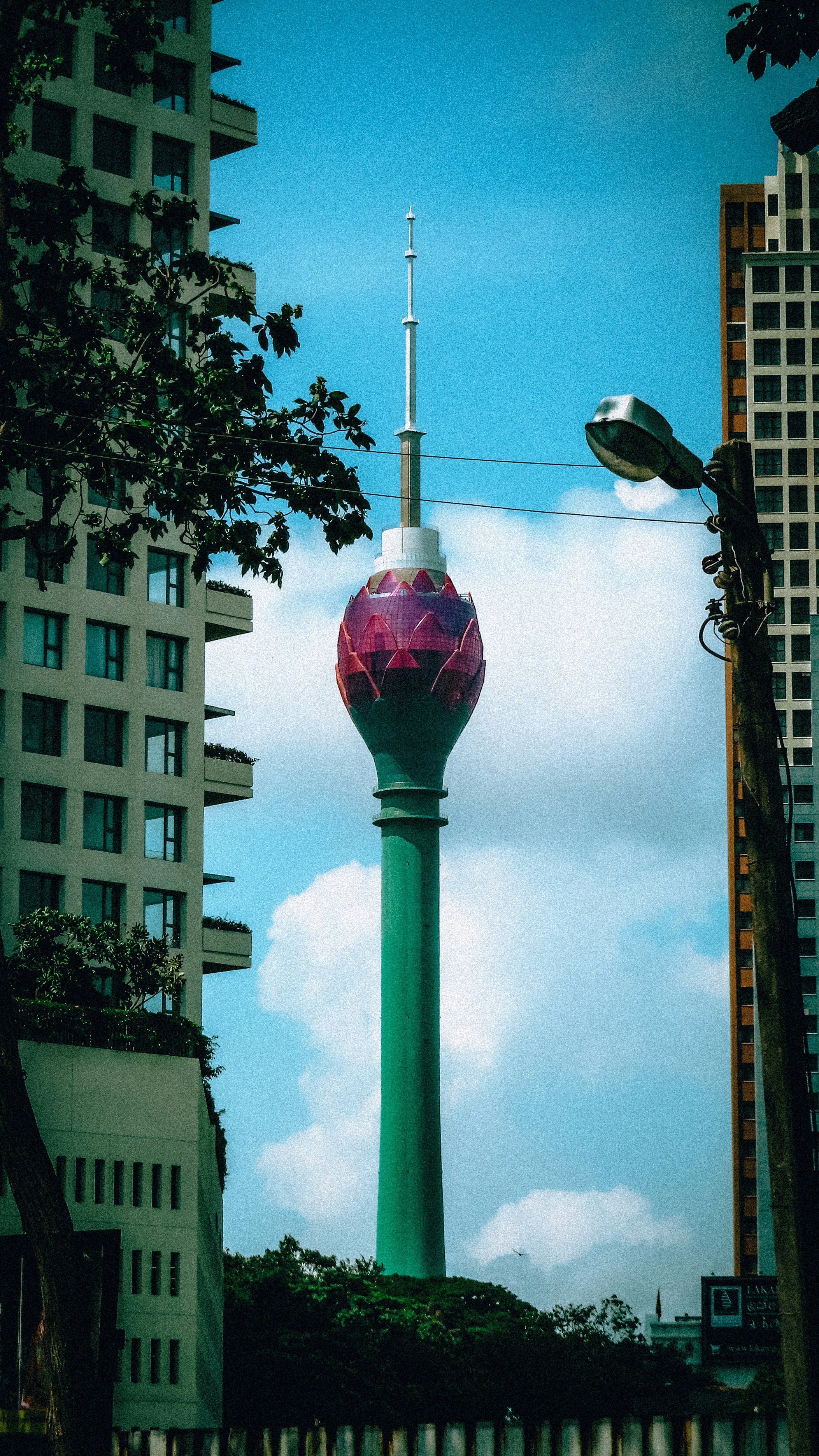 Green observation tower with a purple geometric dome, flanked by modern buildings against a bright blue sky.