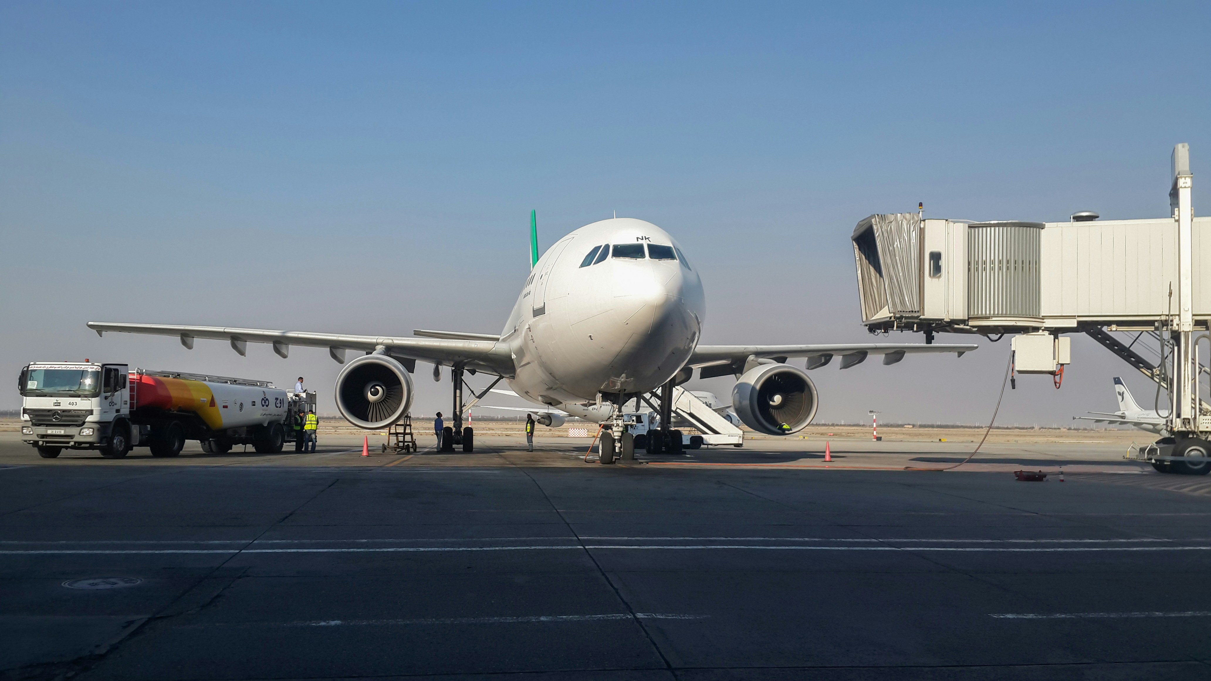 Front-on shot of a commercial airliner docked at a jet bridge, with ground support vehicles visible on the tarmac.