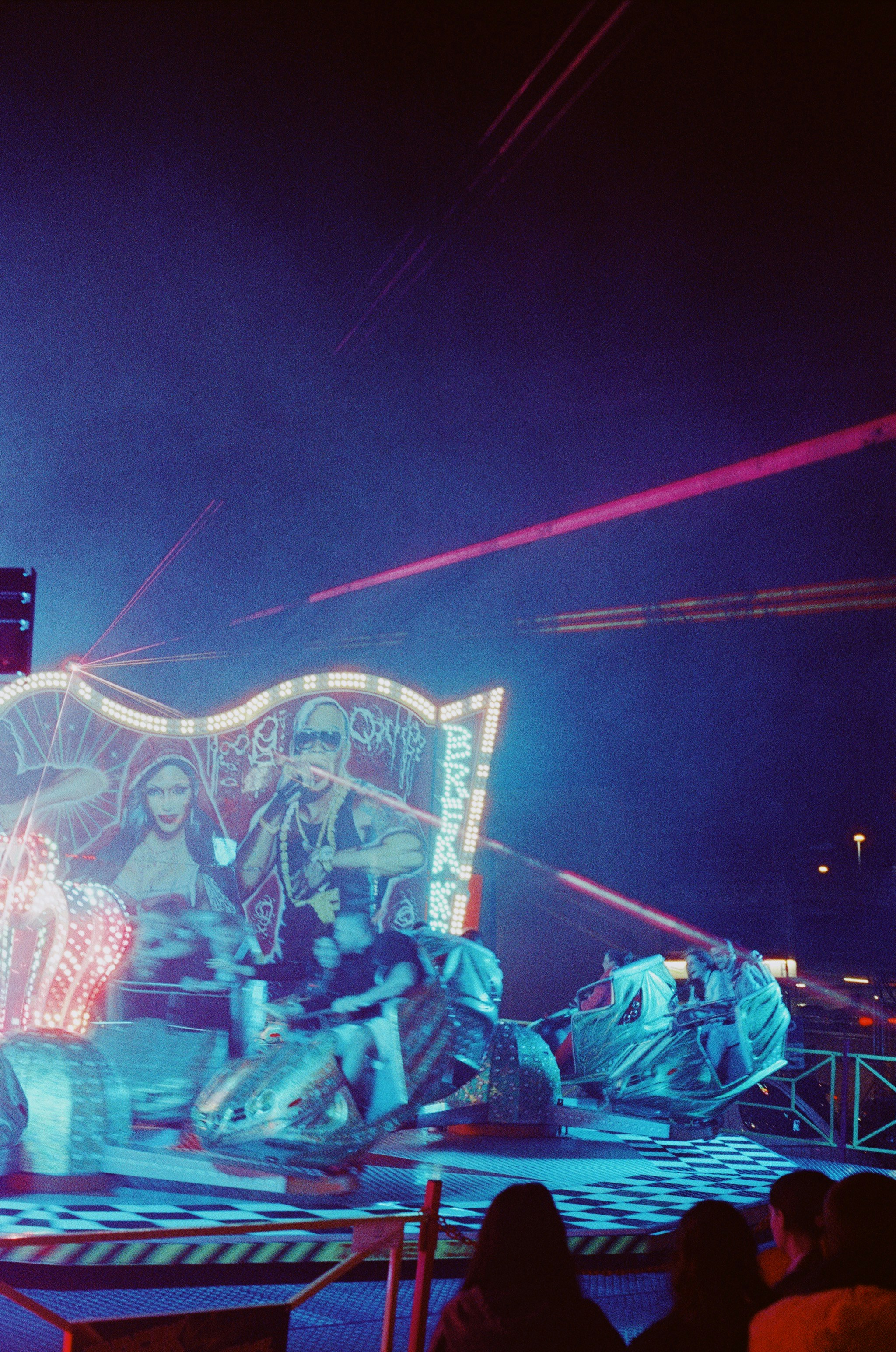 Night photograph of a carnival ride with bright marquee lights and motion blur, spectators in the foreground.
