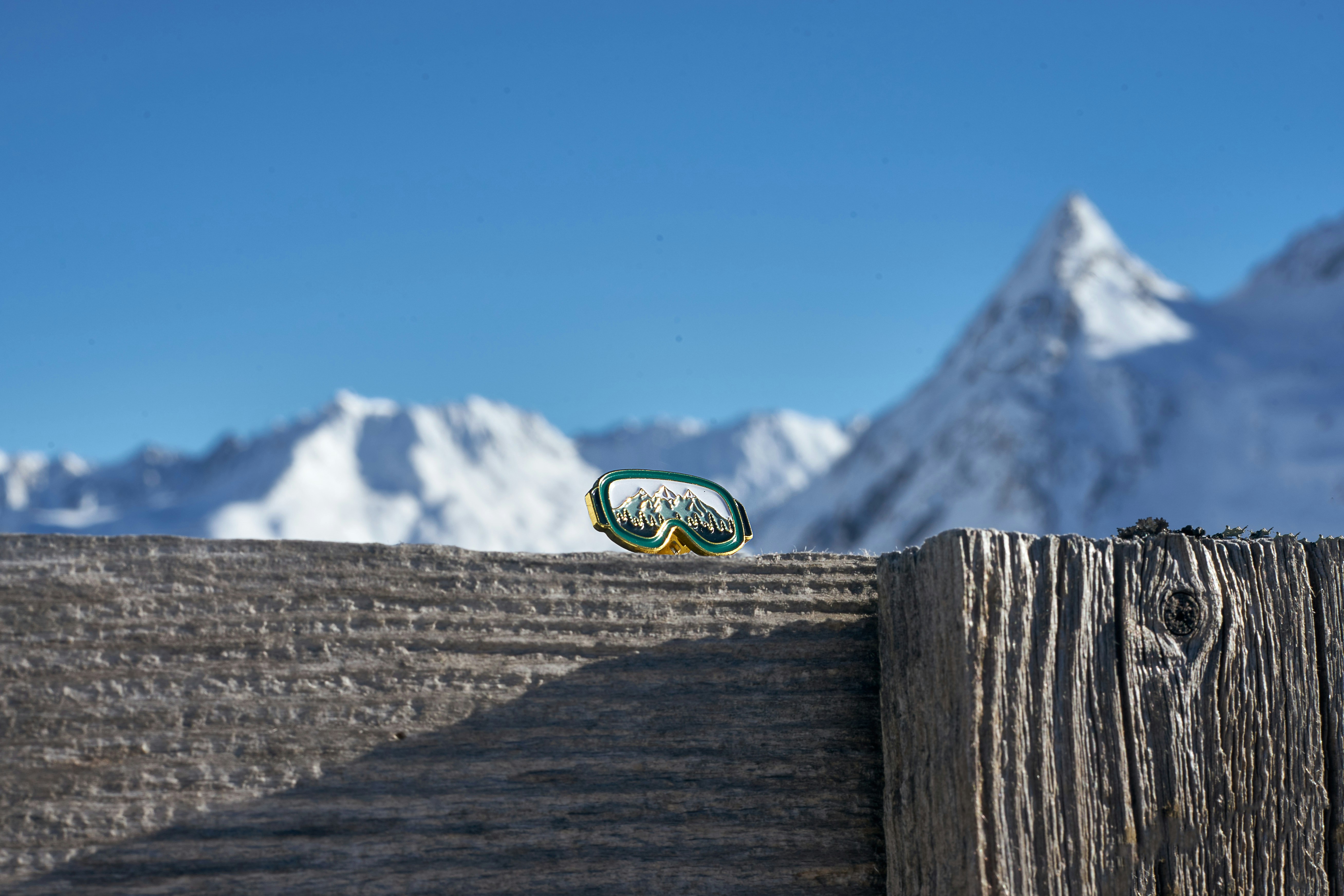 Enamel pin of a skiing goggle on a wooden fence before snowy mountains