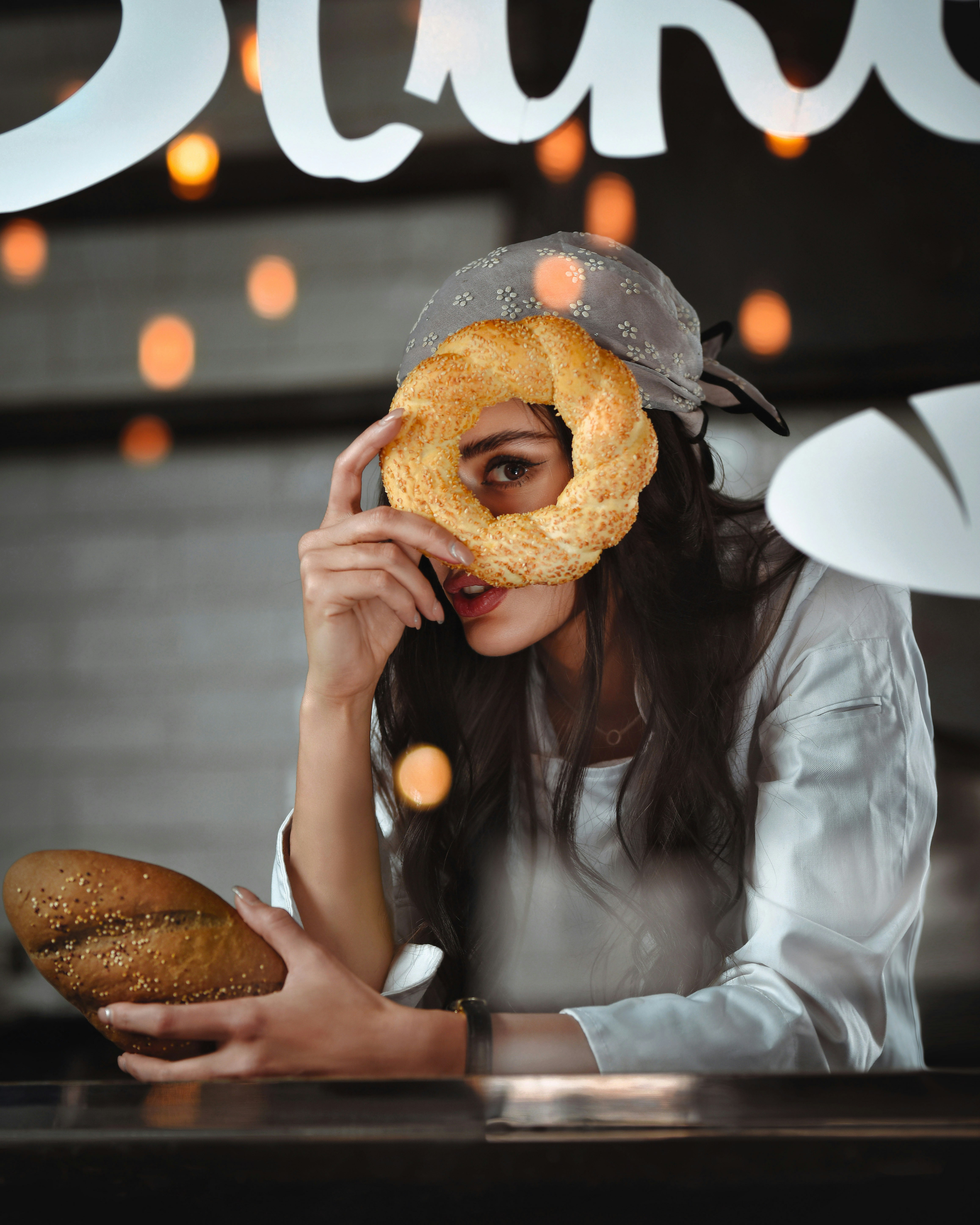Woman playfully looking through a doughnut hole while holding a loaf of bread, surrounded by warm bakery ambiance.