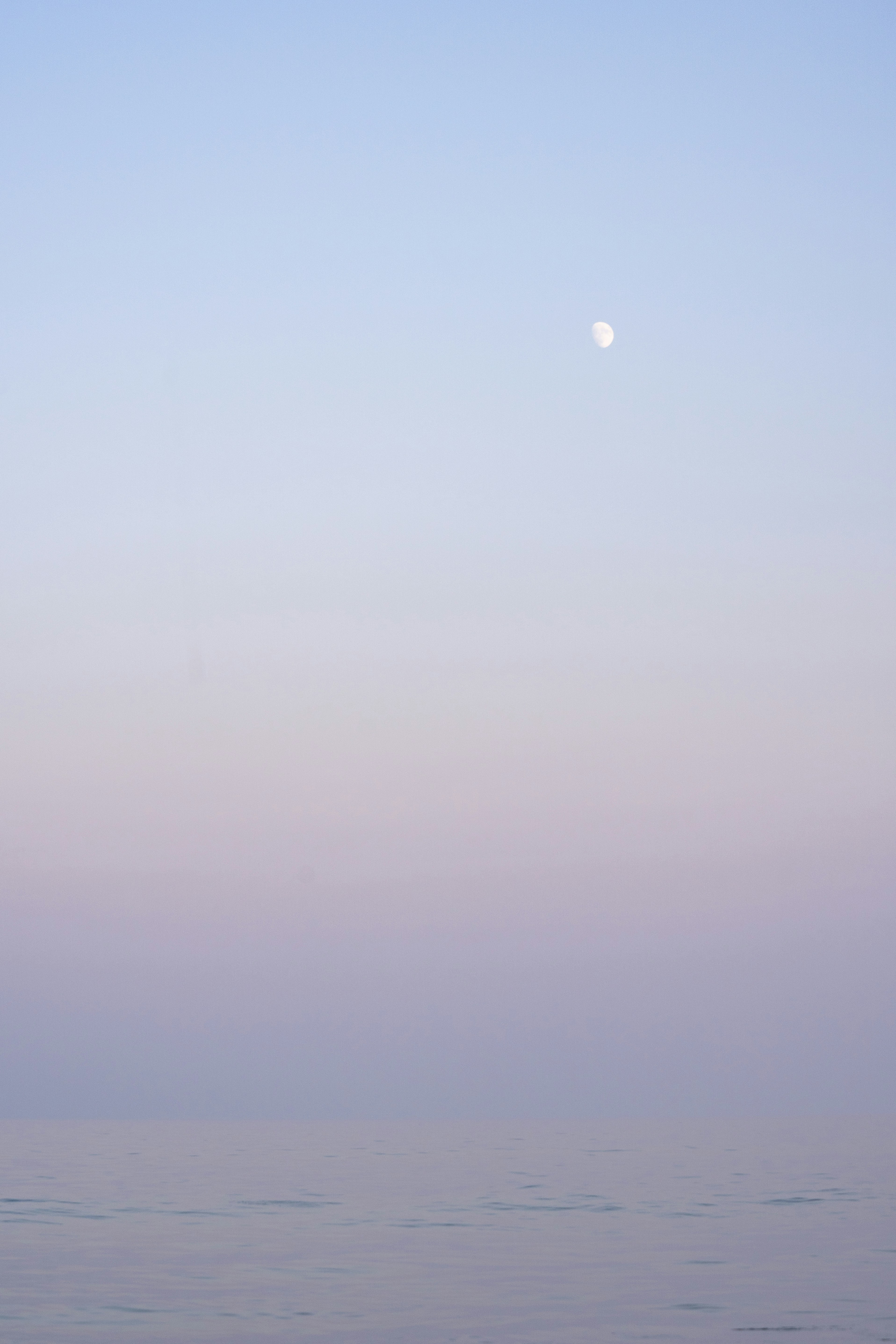 Gentle waves reflect a soft pastel sky at twilight, with a crescent moon hanging above the horizon.