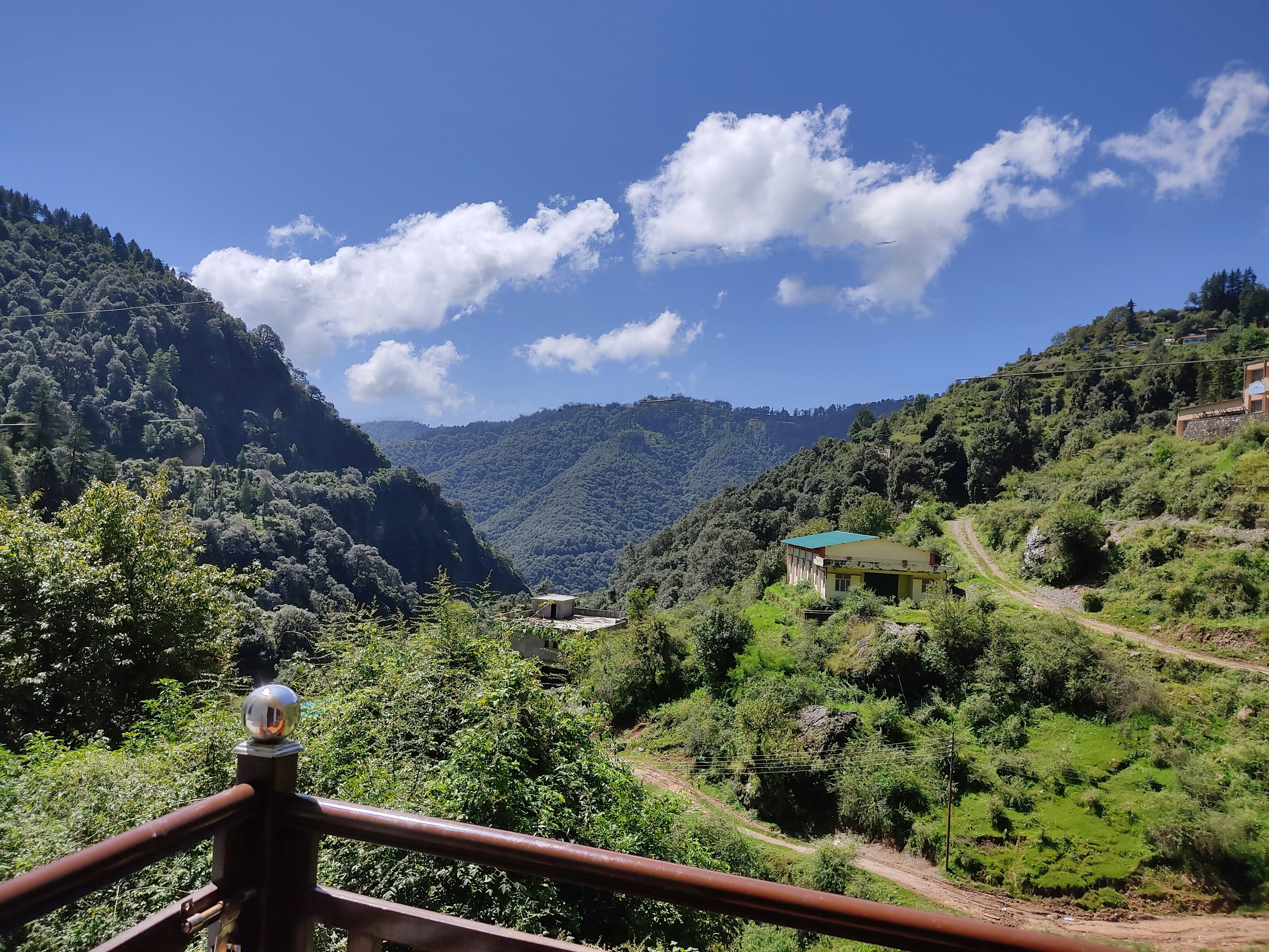 Sunlit terraced hillside landscape with a wooden railing in the foreground and a valley nestled between forested ridges. A green-roofed house sits on the hillside to the right.