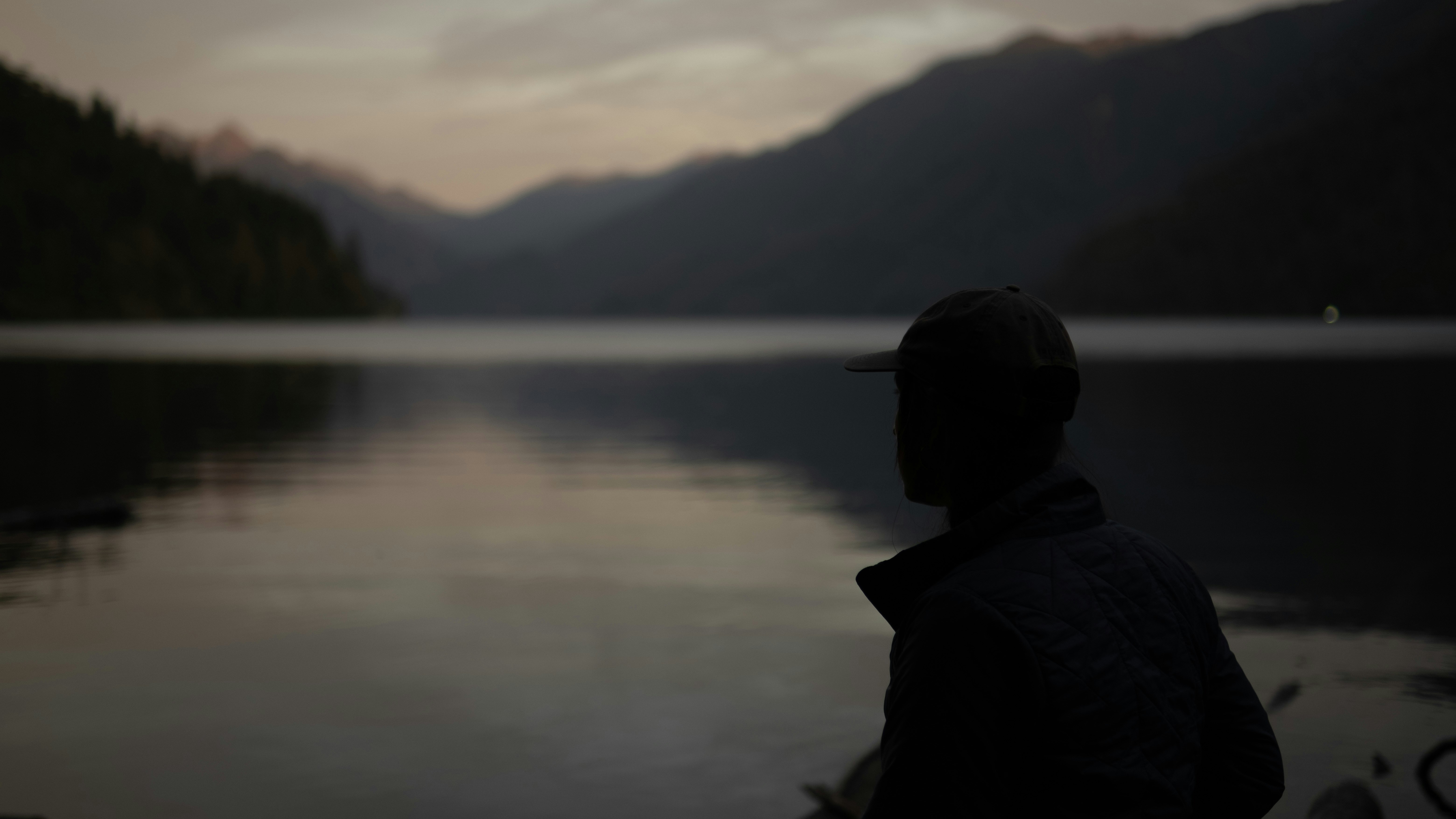 Silhouette of a person gazing across a tranquil lake surrounded by mountains at twilight. The calm water reflects the fading light of the day.