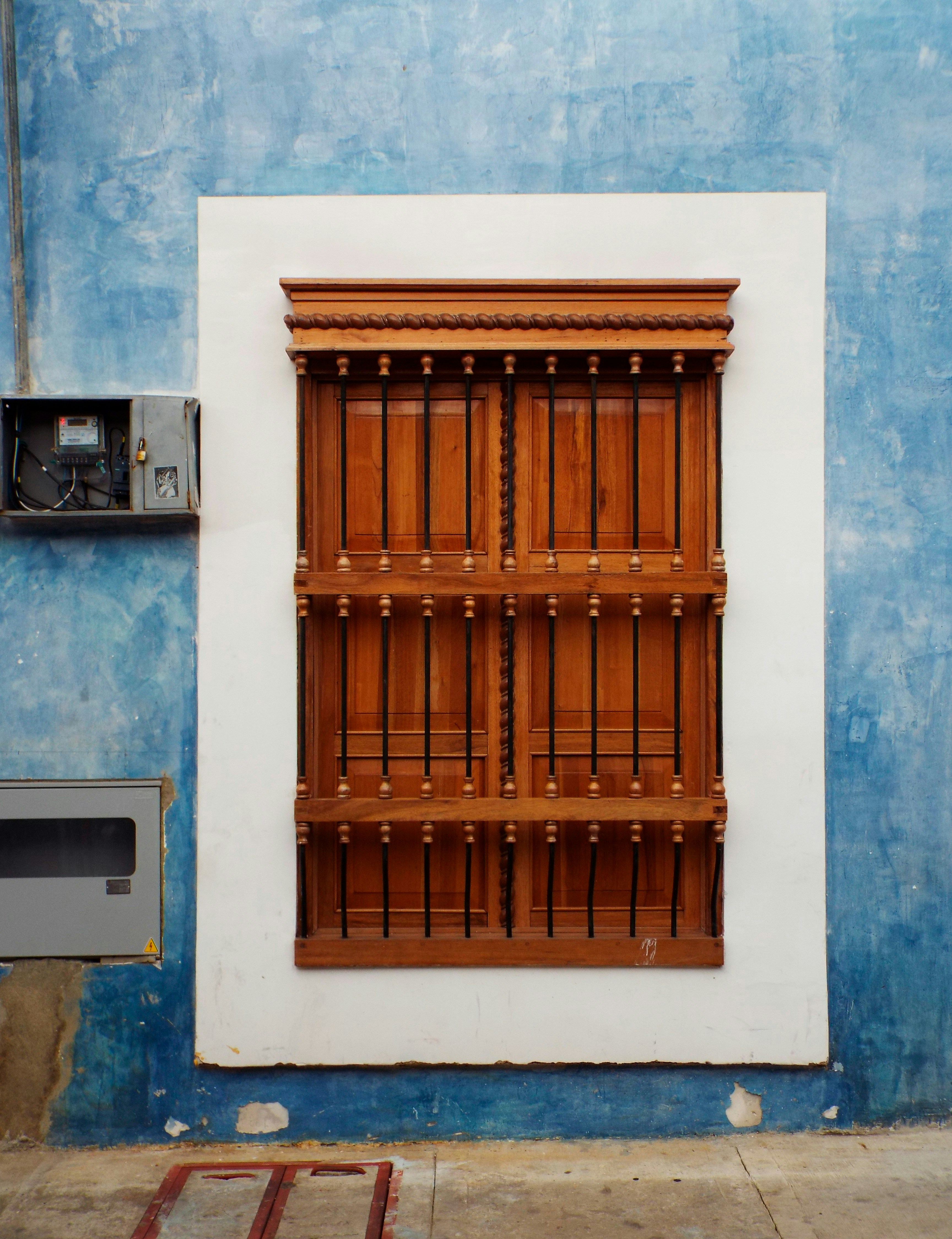 Weathered cobalt wall frames a white-edged wooden window with vertical bars, creating strong architectural contrast.