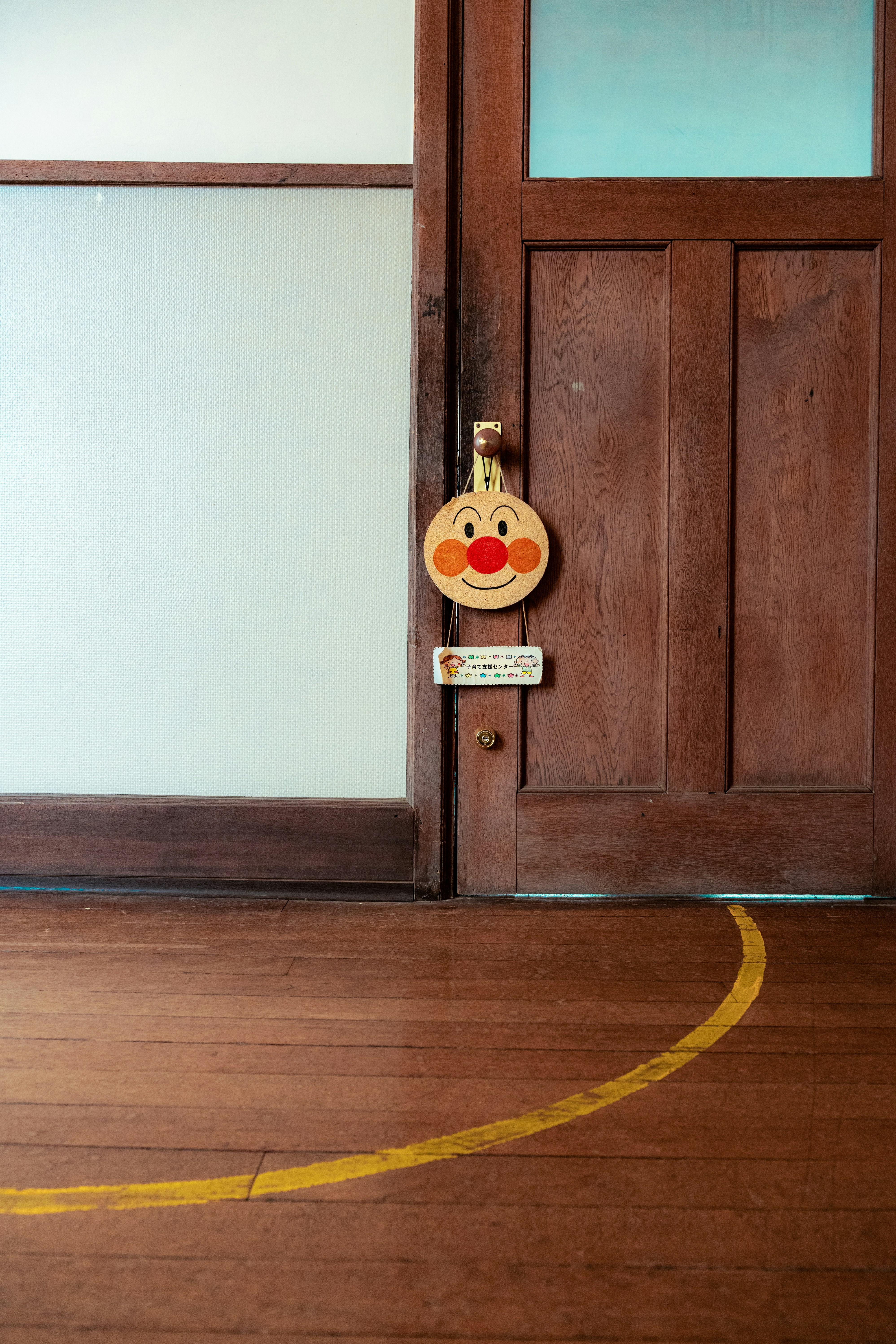 A decorative round wooden face with red cheeks hangs from a door handle on a wooden door. A small sign hangs beneath, adding a charming touch to the doorway.