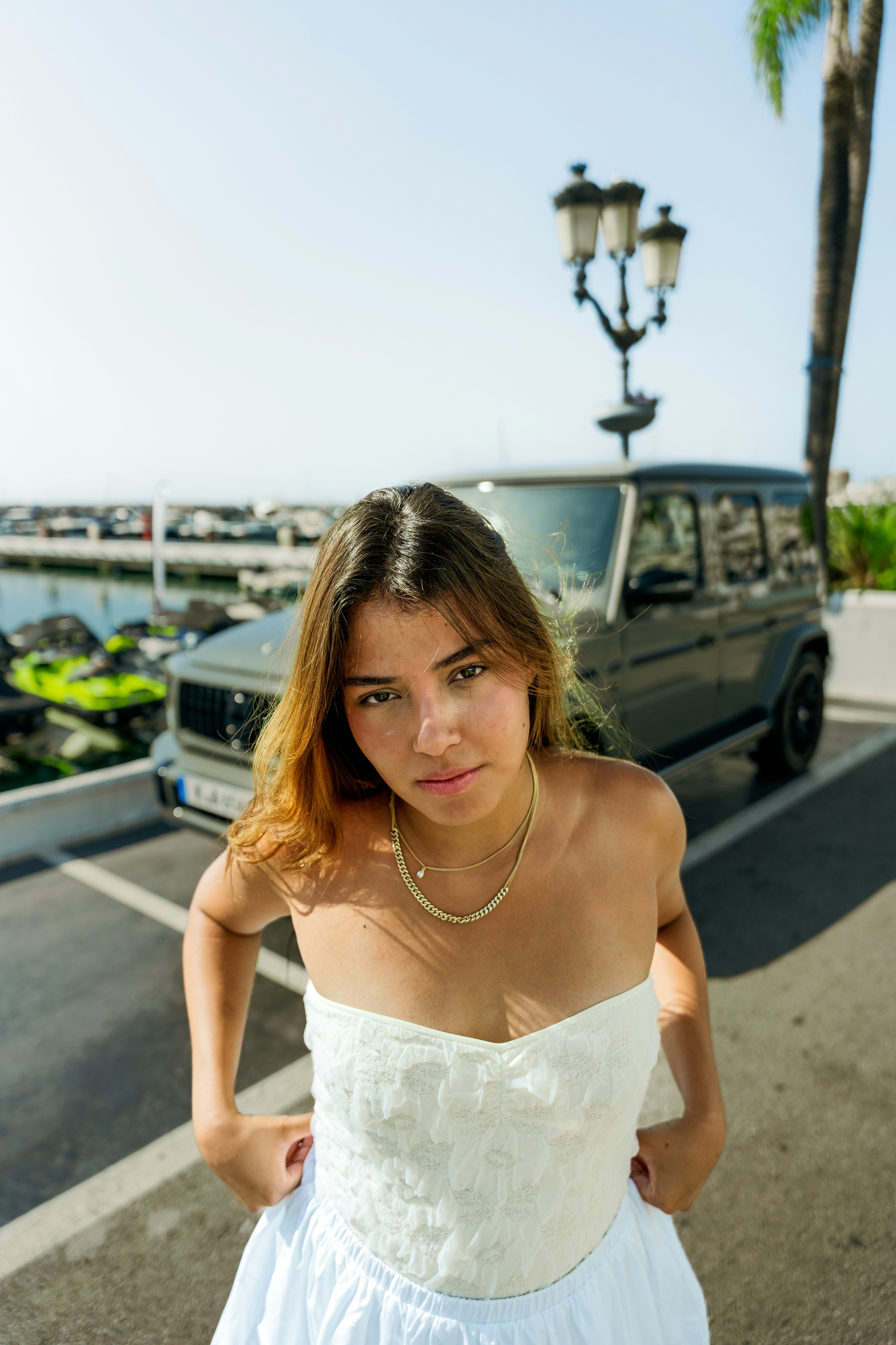 A young woman stands confidently in a sunlit parking area, with a vintage vehicle and marina in the background. Her expression reflects a blend of poise and curiosity.