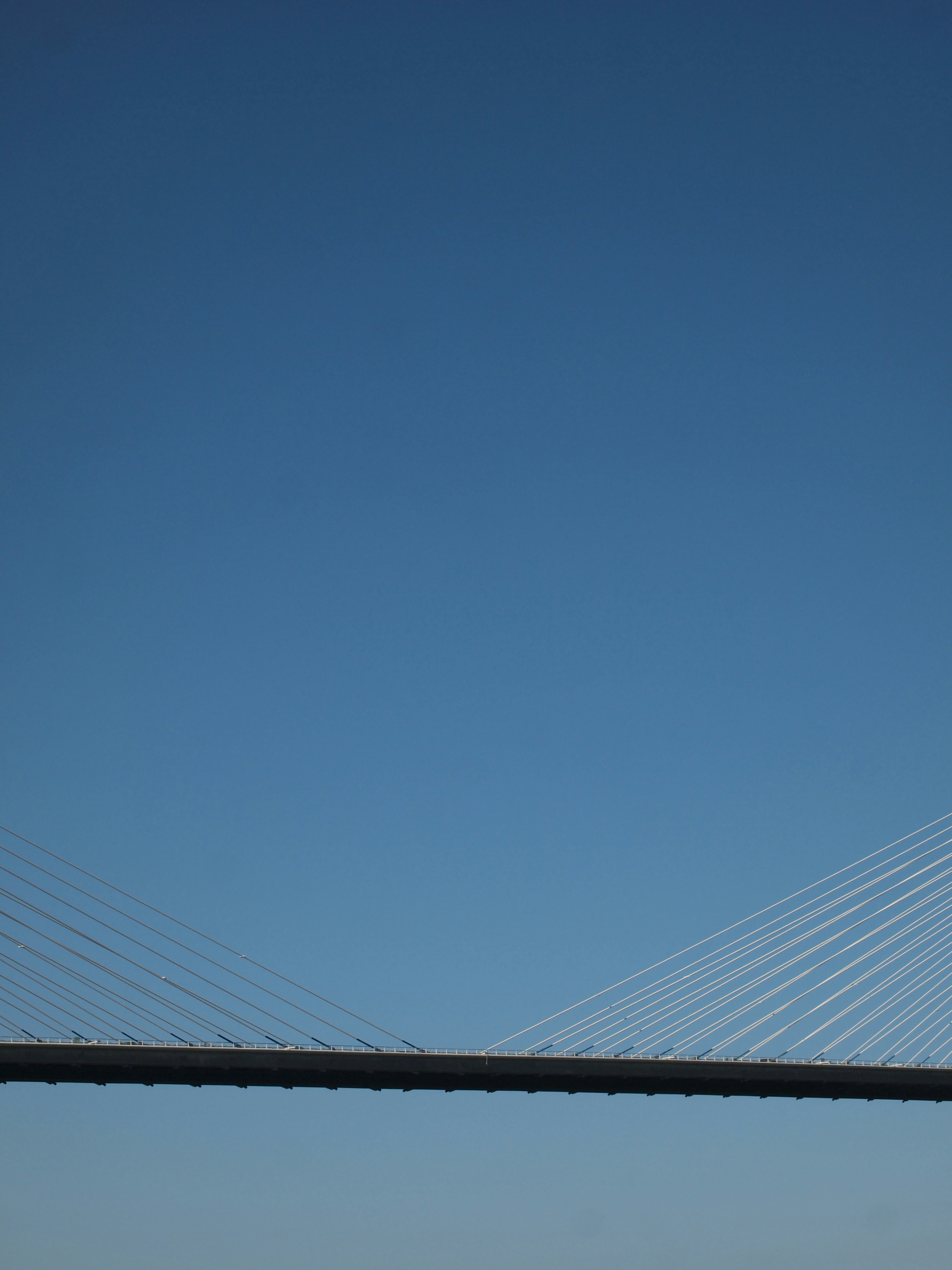 A suspension bridge spans the lower portion of a clear blue sky, its diagonal cables fanning toward the horizon. The composition emphasizes geometric lines and architectural detail.