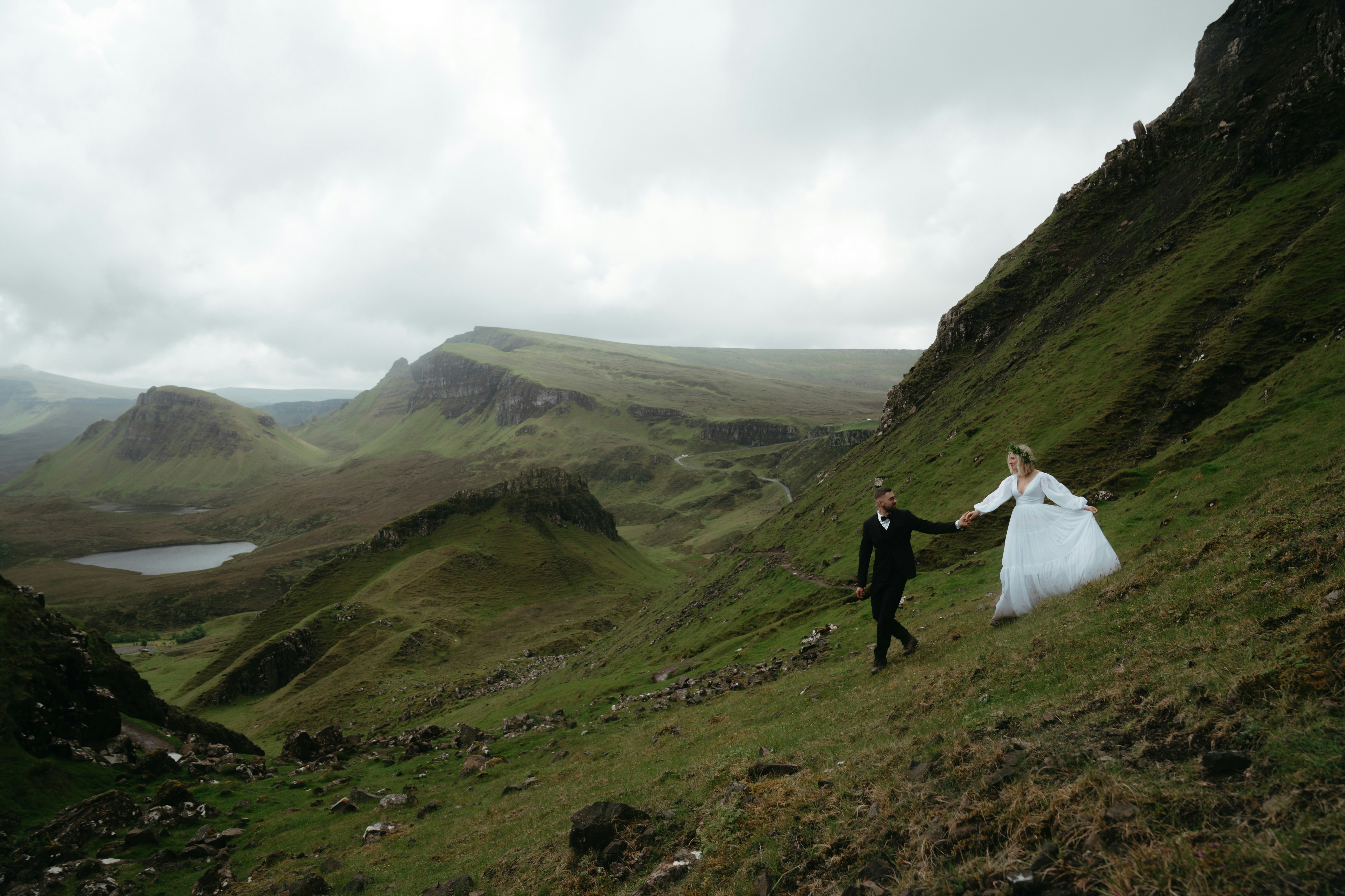 Bride and groom holding hands on a lush hillside in the Isle of Skye under a cloudy sky.