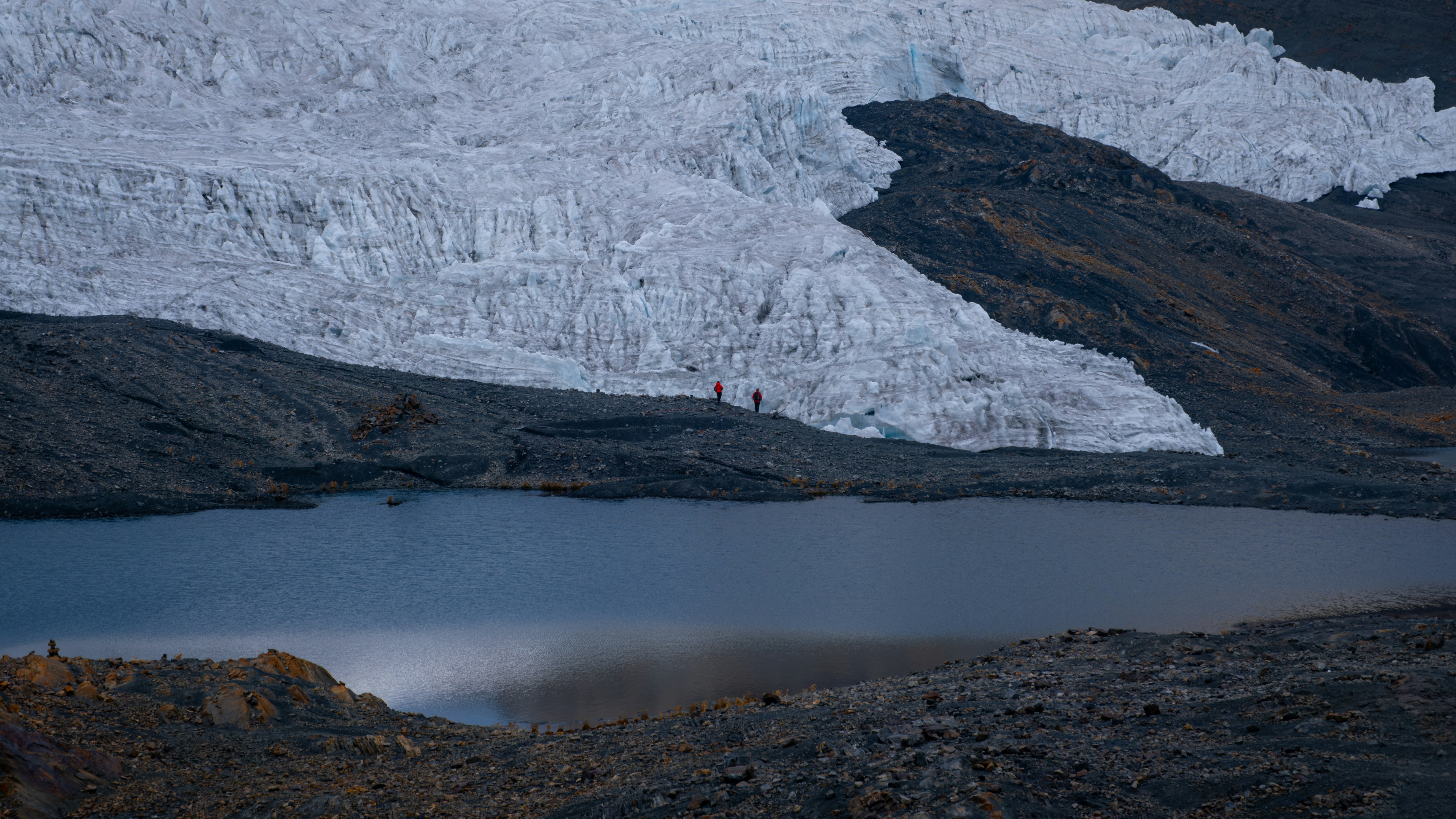 Two figures approach the immense Pastoturi glacier with a serene lake in the foreground.