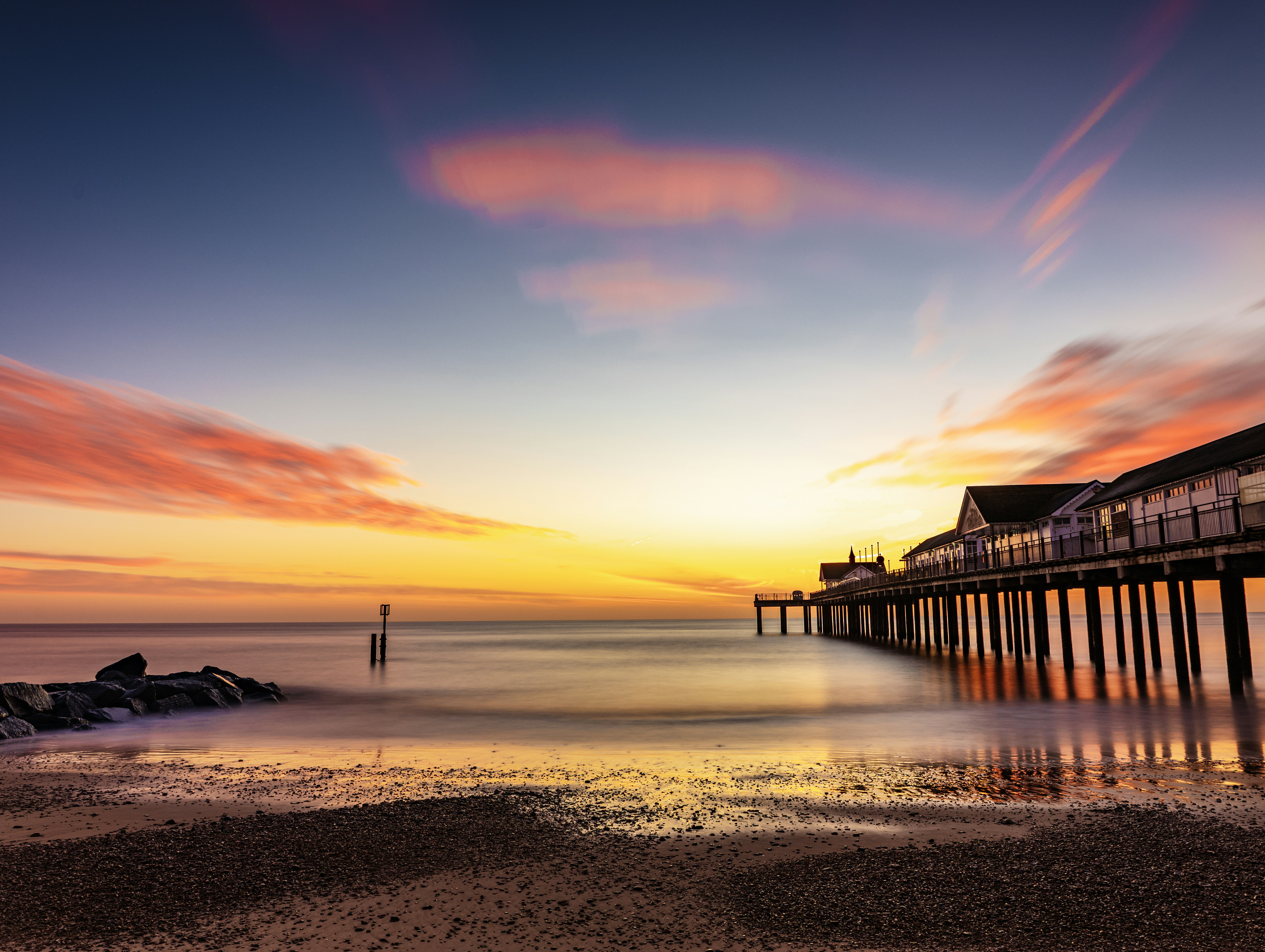 A serene coastal scene featuring a pier extending into a calm sea under a vibrant sunset sky with wispy clouds.