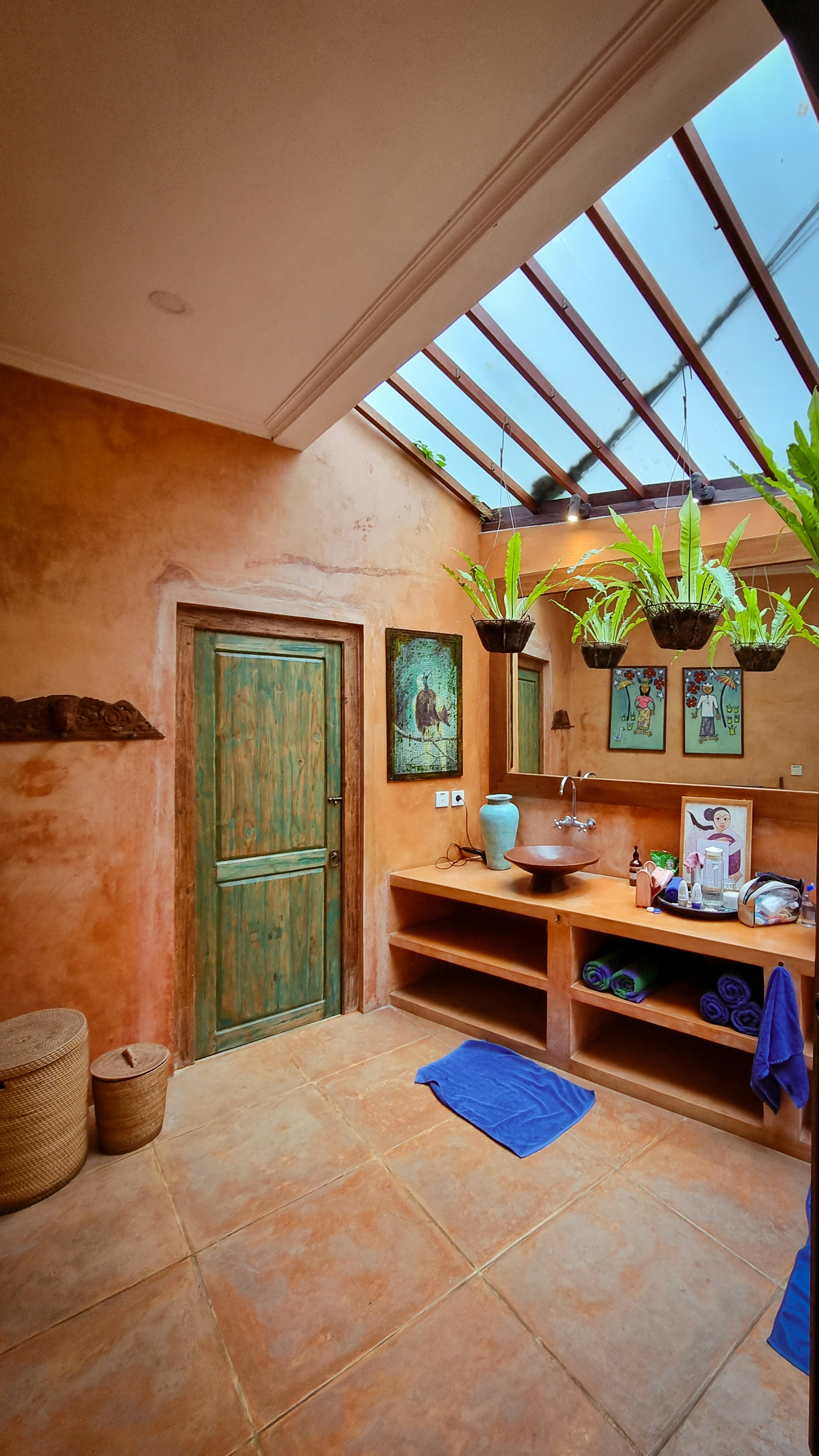 Terracotta walls frame a skylit bathroom with hanging plants above a wooden vanity and a weathered green door. Warm, natural light highlights textures, decor, and towels, giving the space a sunlit, earthy ambiance.