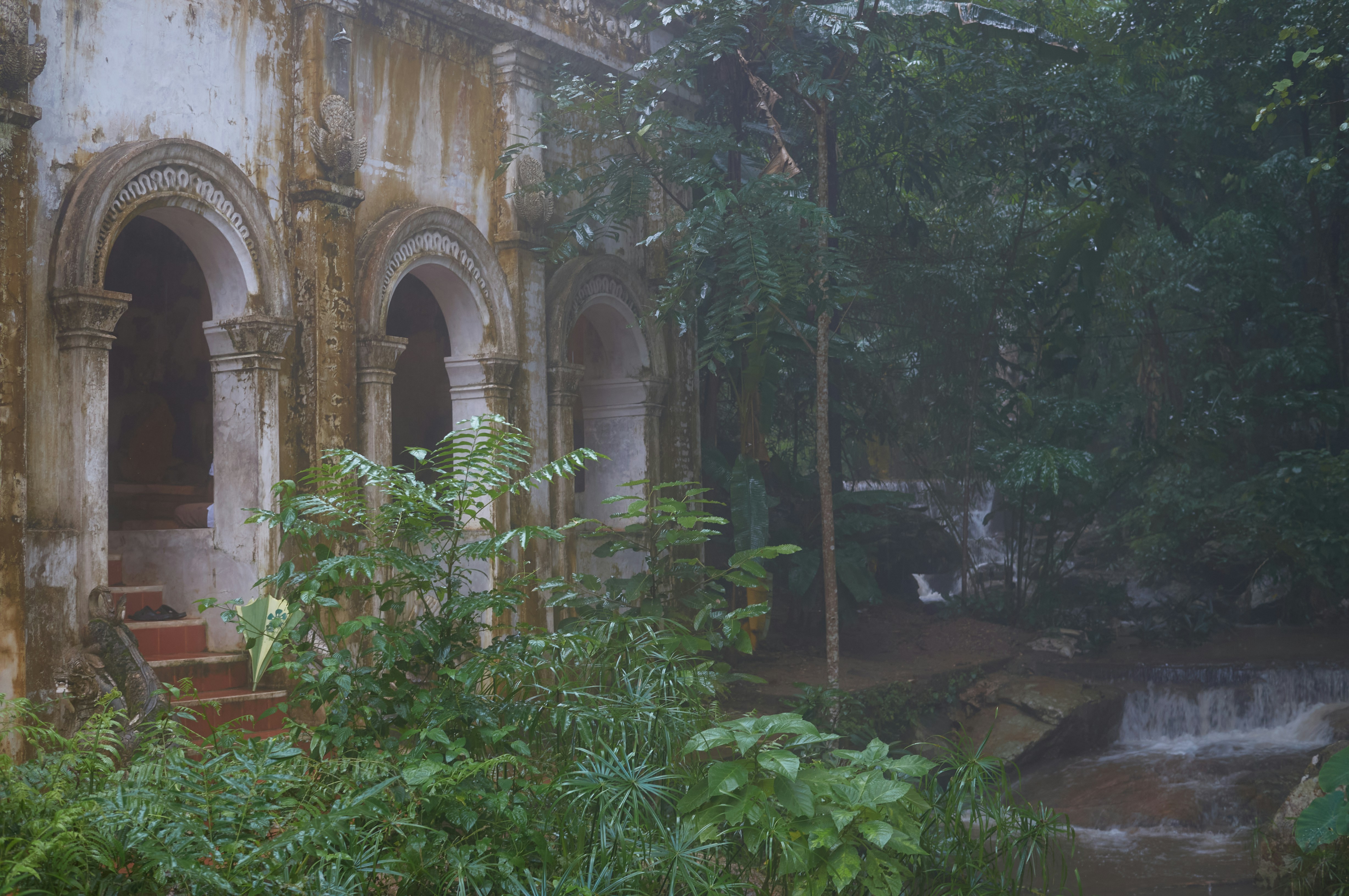 Stone arches overgrown with plants frame a secluded ruin beside a forest stream, blending architectural detail with lush greenery.