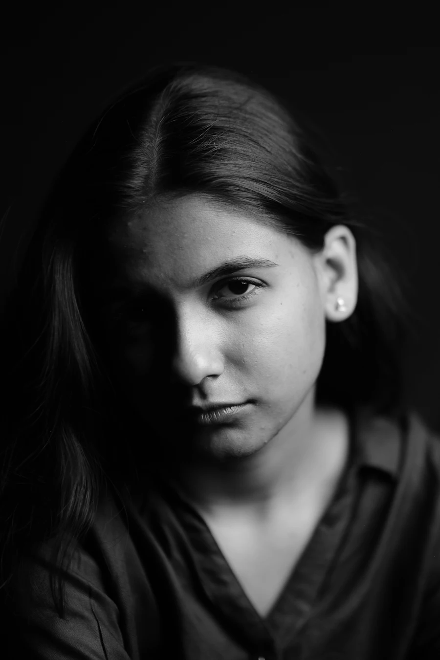 Black-and-white portrait of a young woman against a dark background, lit dramatically from one side. Her face and eye are the clear focal point, with hair softly framing the composition.