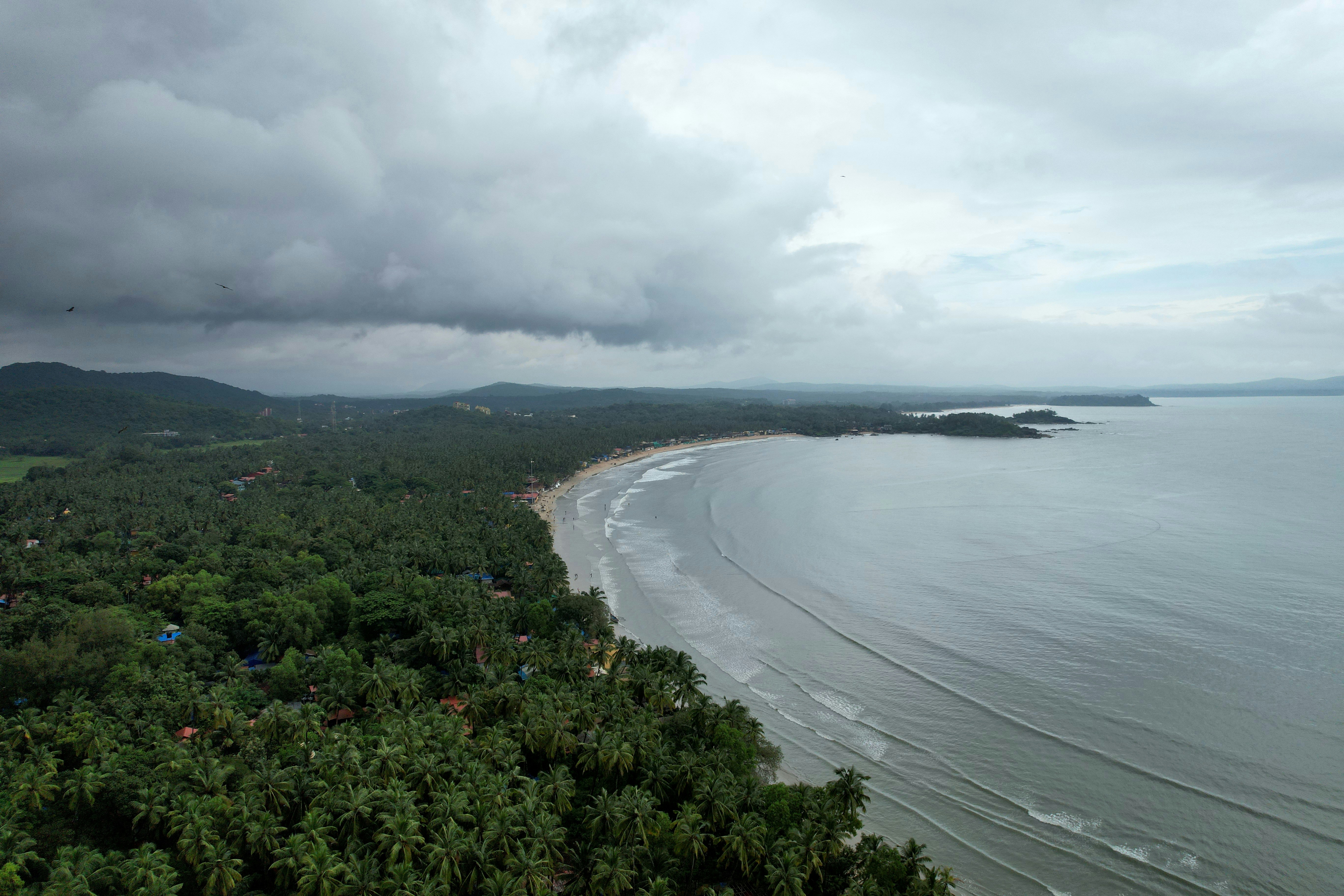 Palolem Beach is a stretch of white sand on a bay in Goa, South India. It's known for its calm waters. Lined with palm trees, fishing boats, and colorful wooden shacks, the beach faces Canacona Island, known for its resident monkeys. | An aerial view of a beach and a body of water