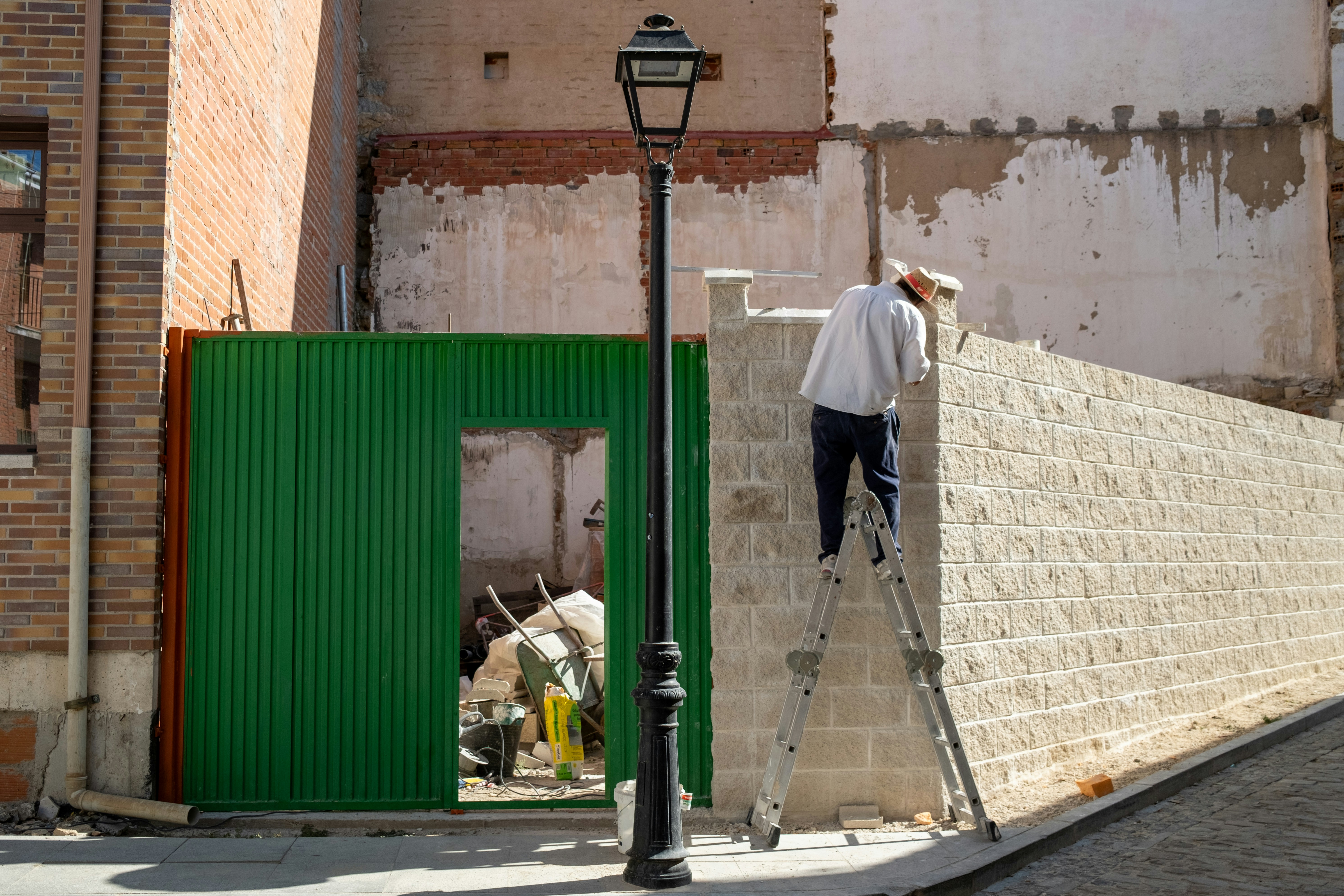 Worker on a ladder building a brick wall beside a green gate in bright sunlight.