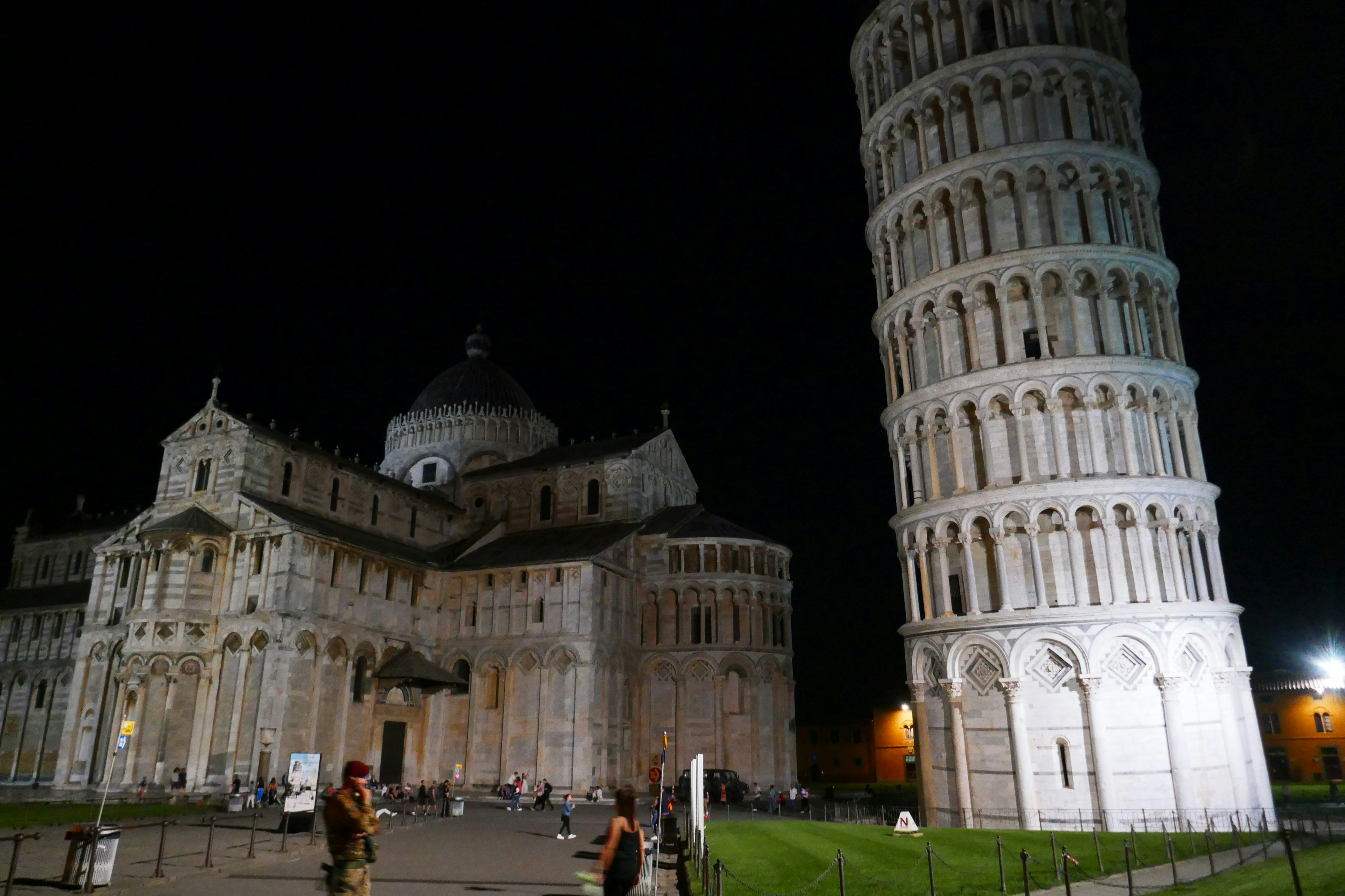 Night view of Pisa's Cathedral and the Leaning Tower illuminated in warm lighting, with visitors strolling the square. A documentary-style photograph captures architectural details and atmospheric glow.