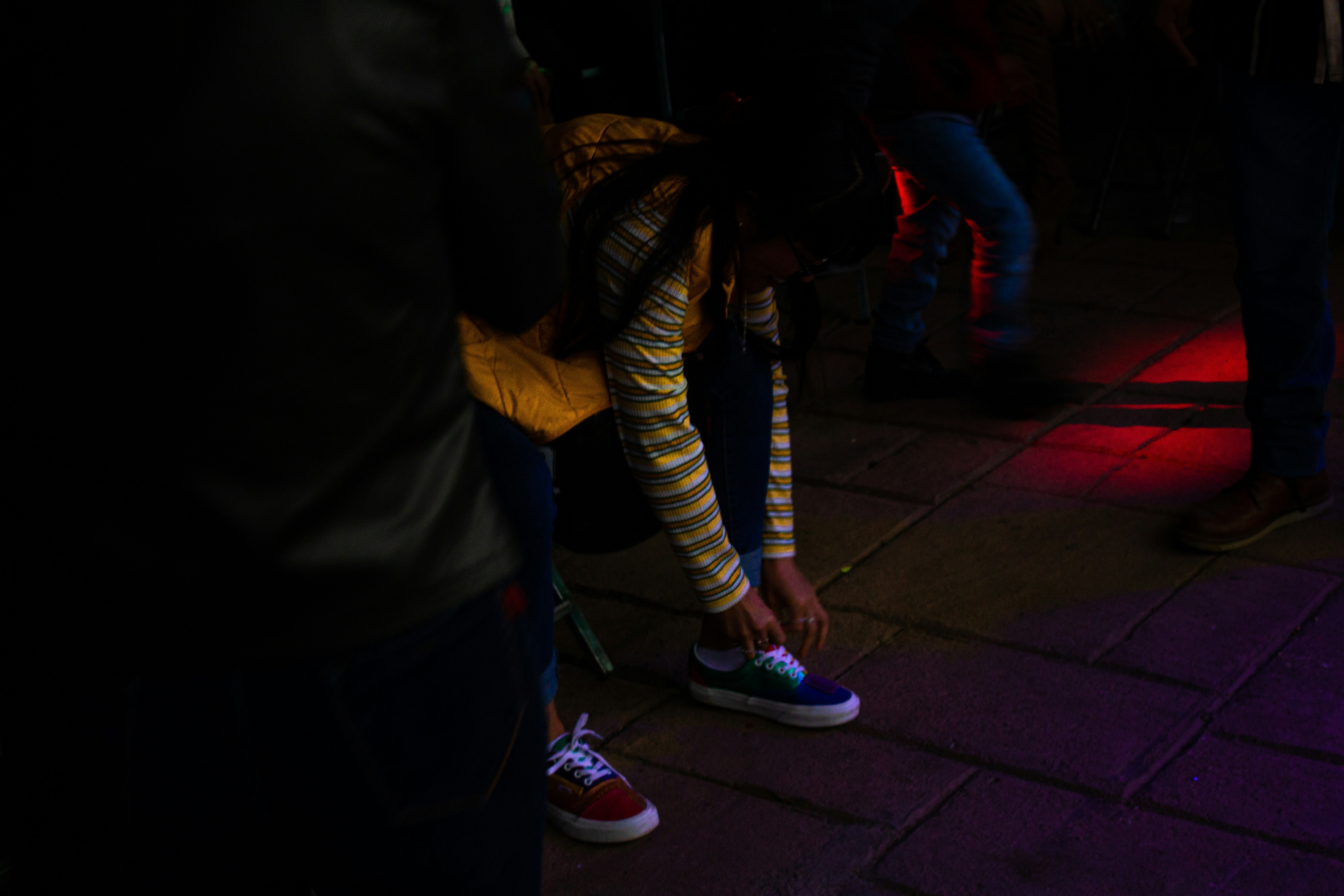 A person kneeling to tie their colorful shoes on a dimly lit street, surrounded by shadows and vibrant lighting effects.
