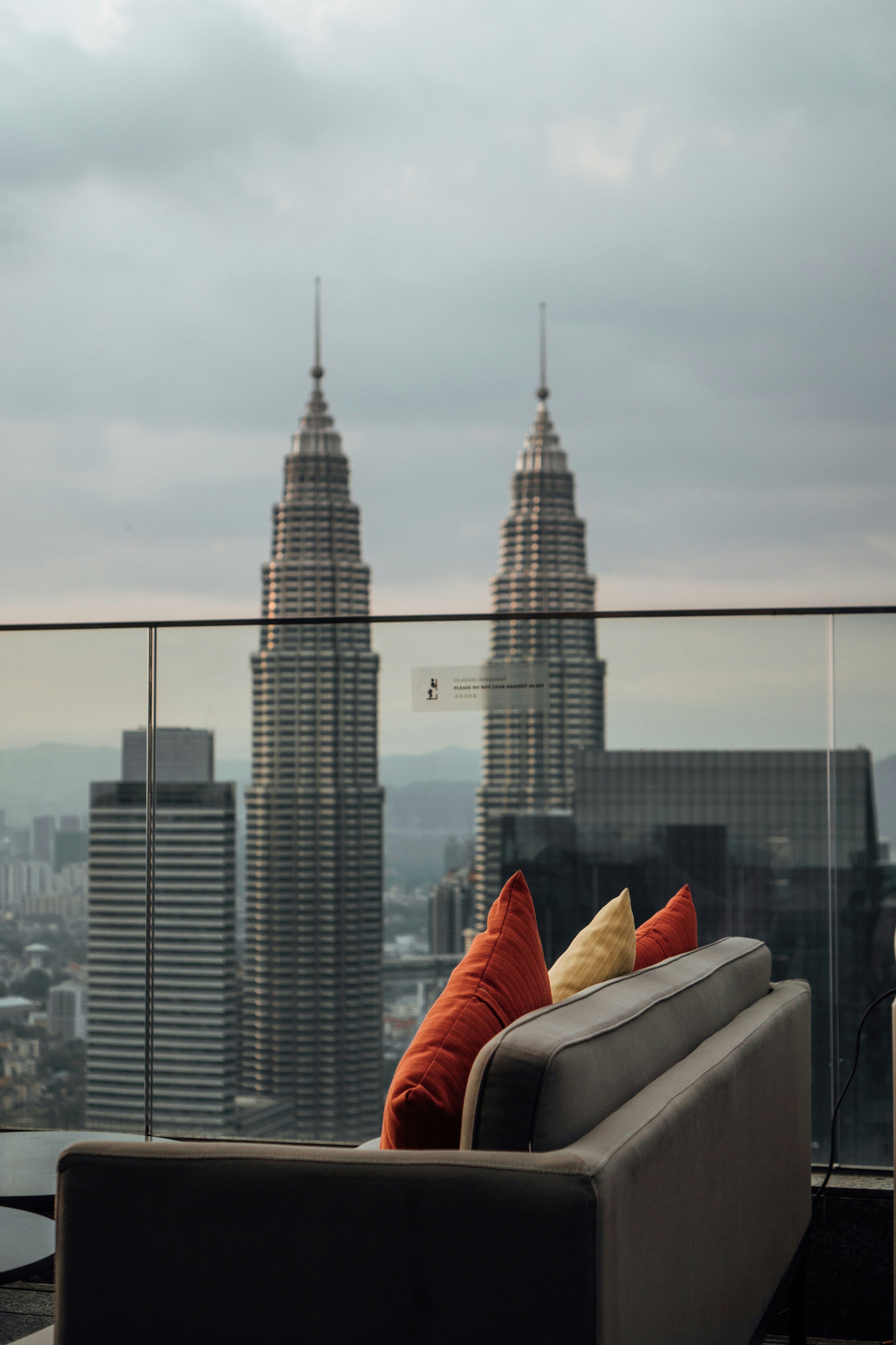 A rooftop lounge with a glass railing and plush red and yellow cushions frames the cityscape. The Petronas Towers rise in the distance, anchoring the skyline.