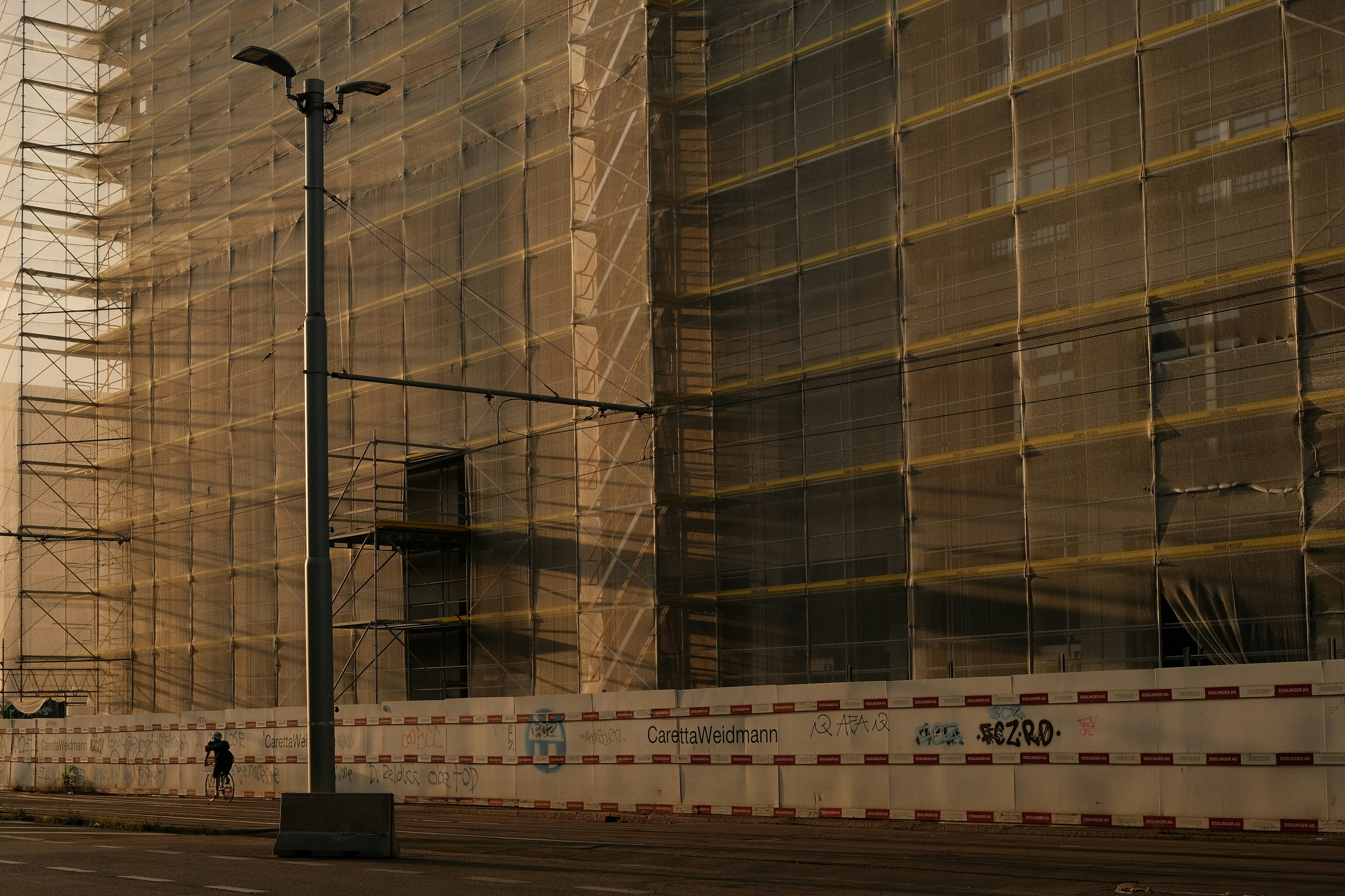Photograph of a city construction site with a tall streetlamp in the foreground and barrier tape along the street, while two figures walk in the distance.