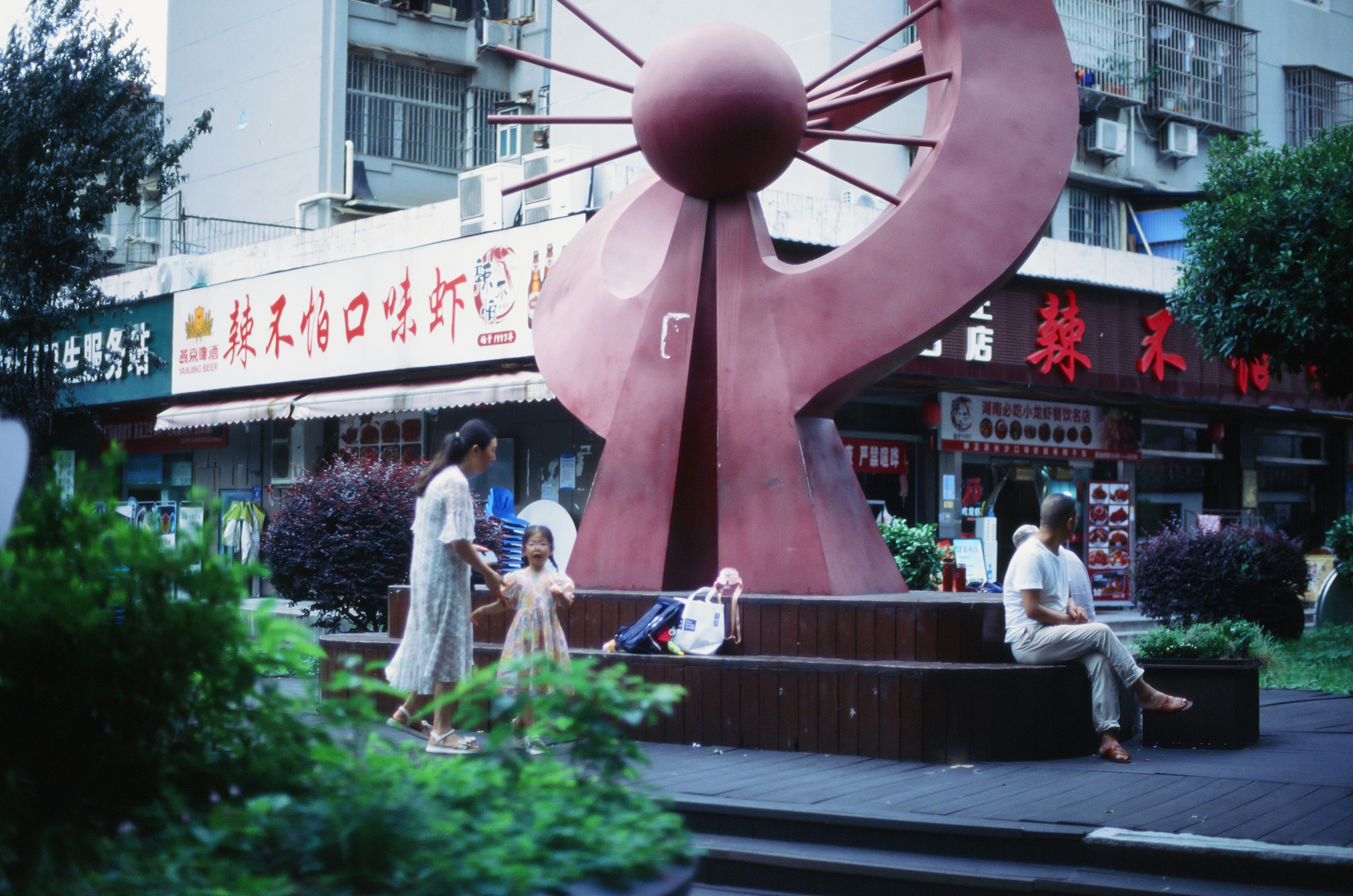 A red sculptural centerpiece dominates a city plaza while passersby pause nearby. Storefronts with Chinese signage frame the scene.