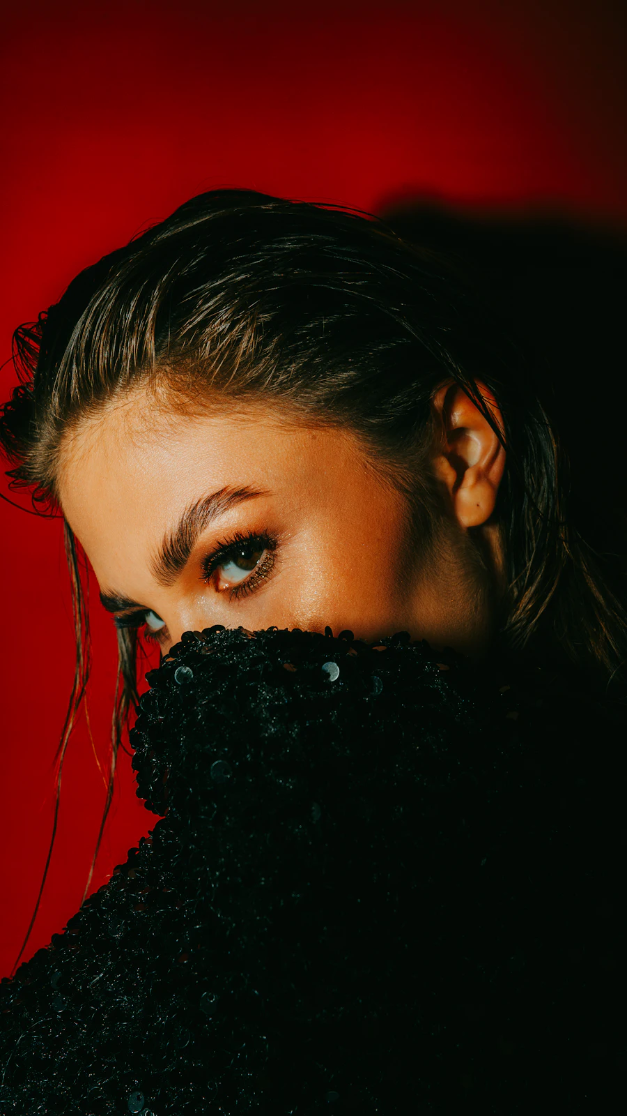 Close-up portrait of a woman with slicked-back hair, one eye sharply visible, and a black sequined garment covering her mouth against a vivid red background.