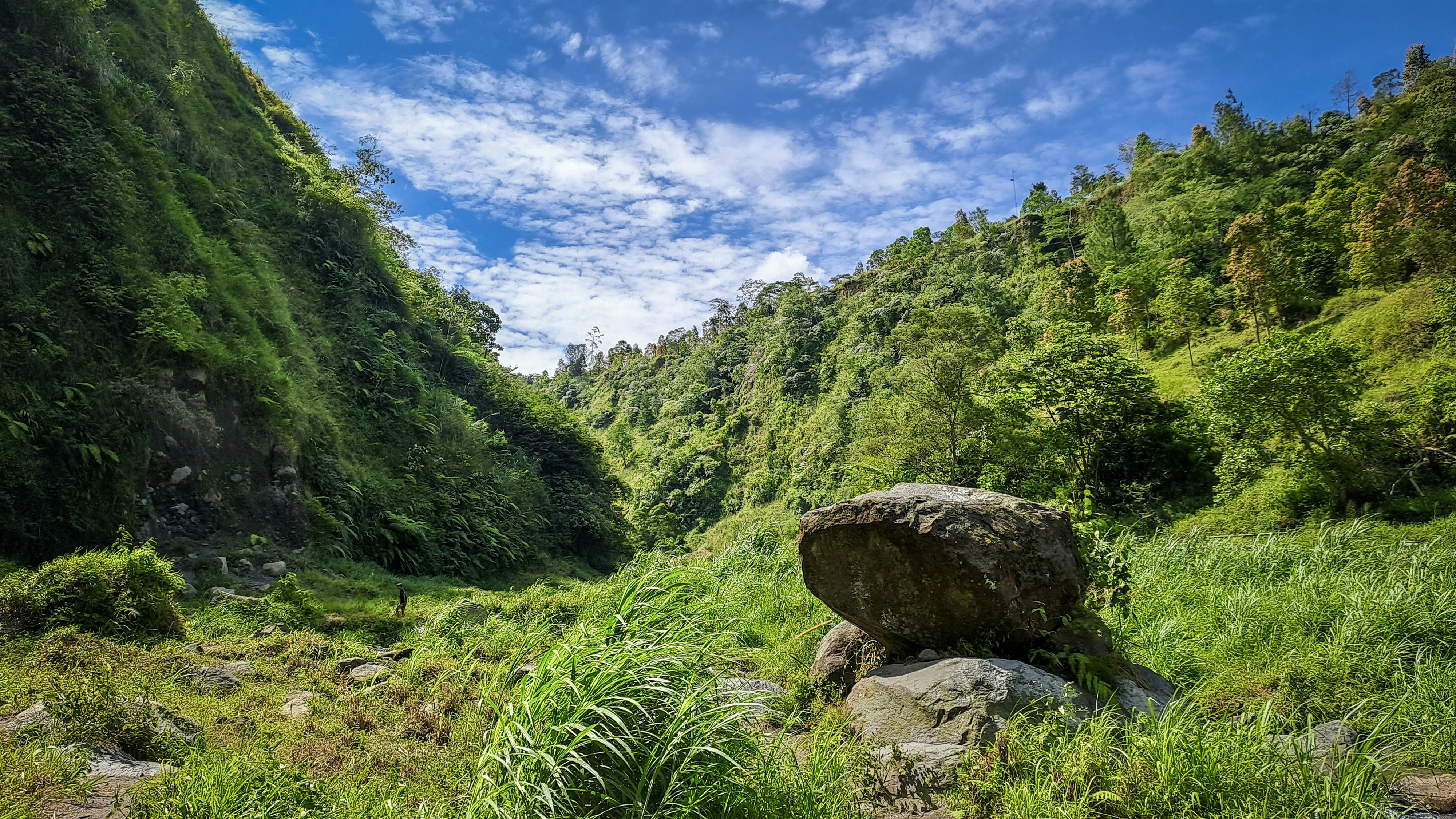 Sunlit valley photograph featuring a prominent rock in the foreground, lush green slopes, and a blue sky.
